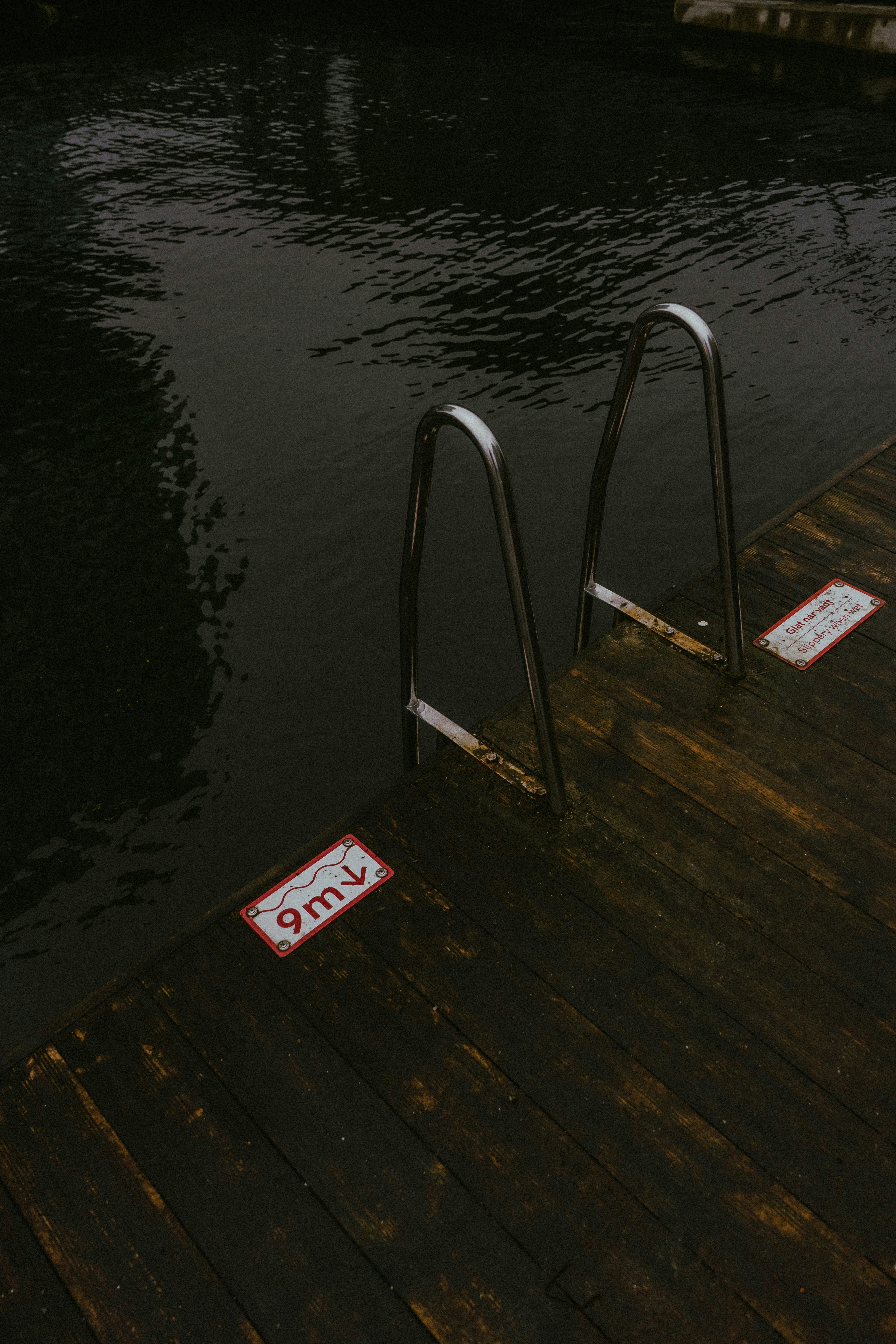 Metal ladder leading into dark water beside a wooden dock, with warning signs partially visible. The surface reflects muted light.