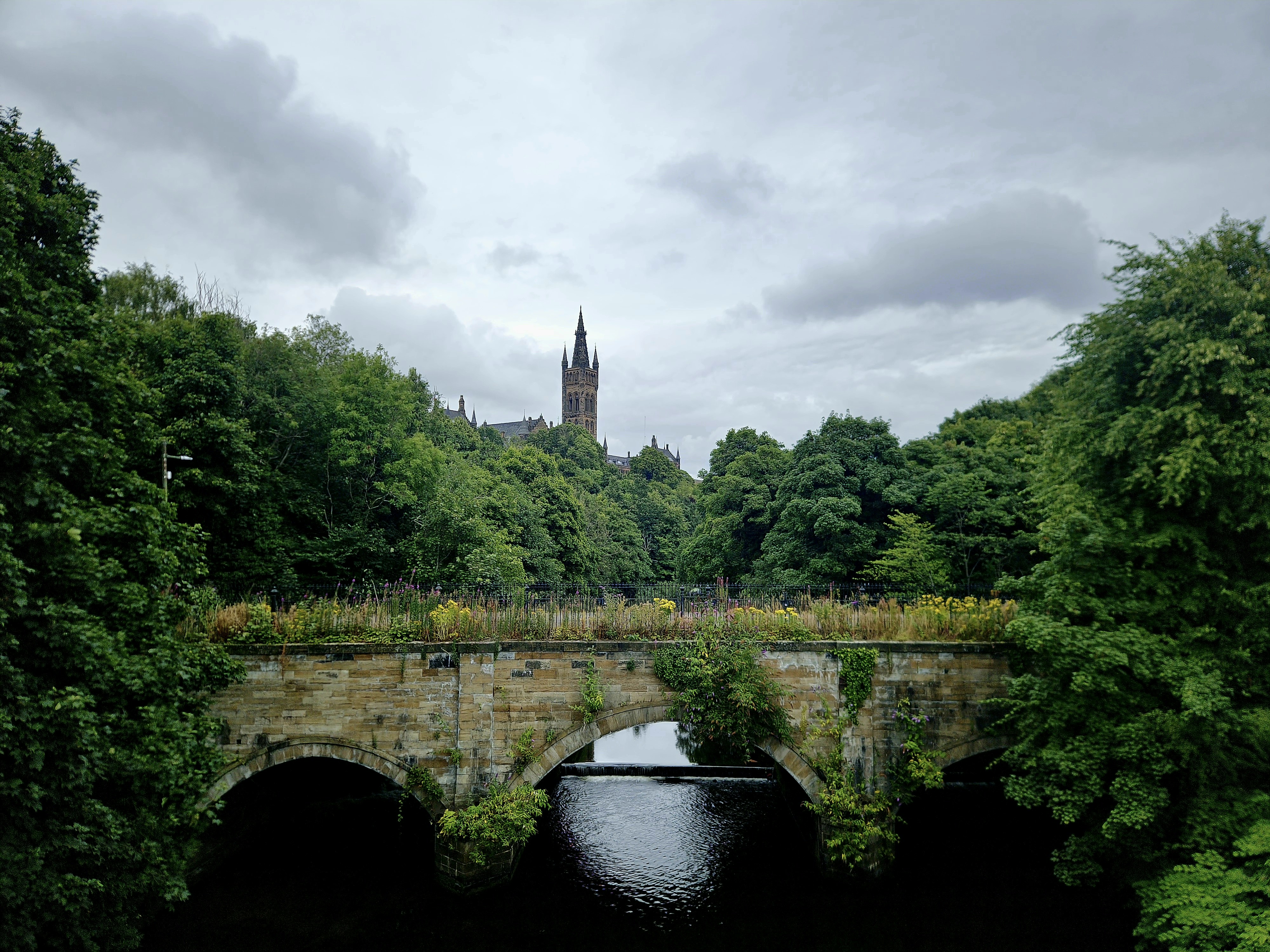 Stone bridge spans a reflective river with lush greenery and a distant spire under a cloudy sky.