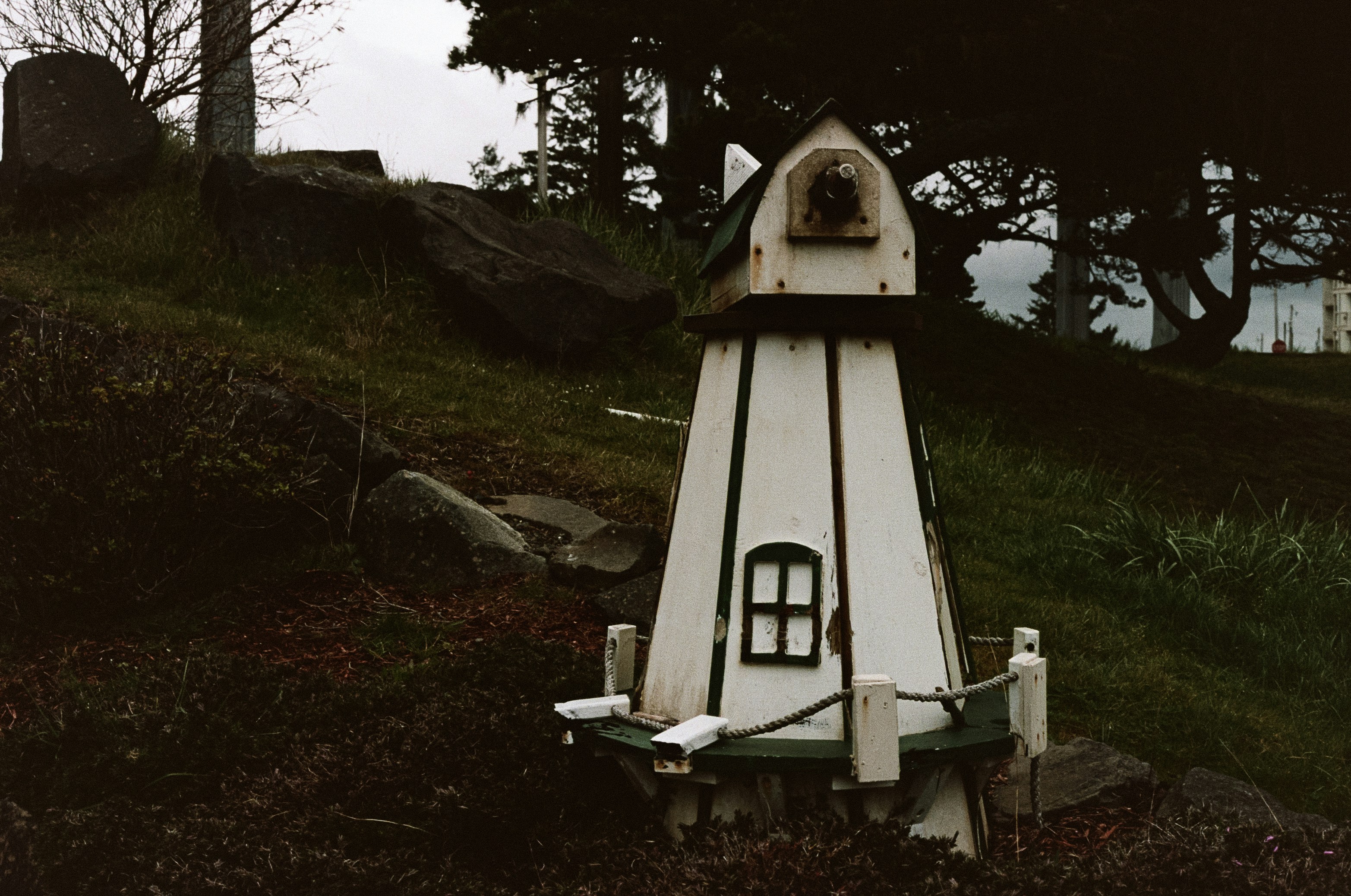 A small white wooden beacon sits on a raised platform on a grassy hill, with pine trees and rocks in the background.