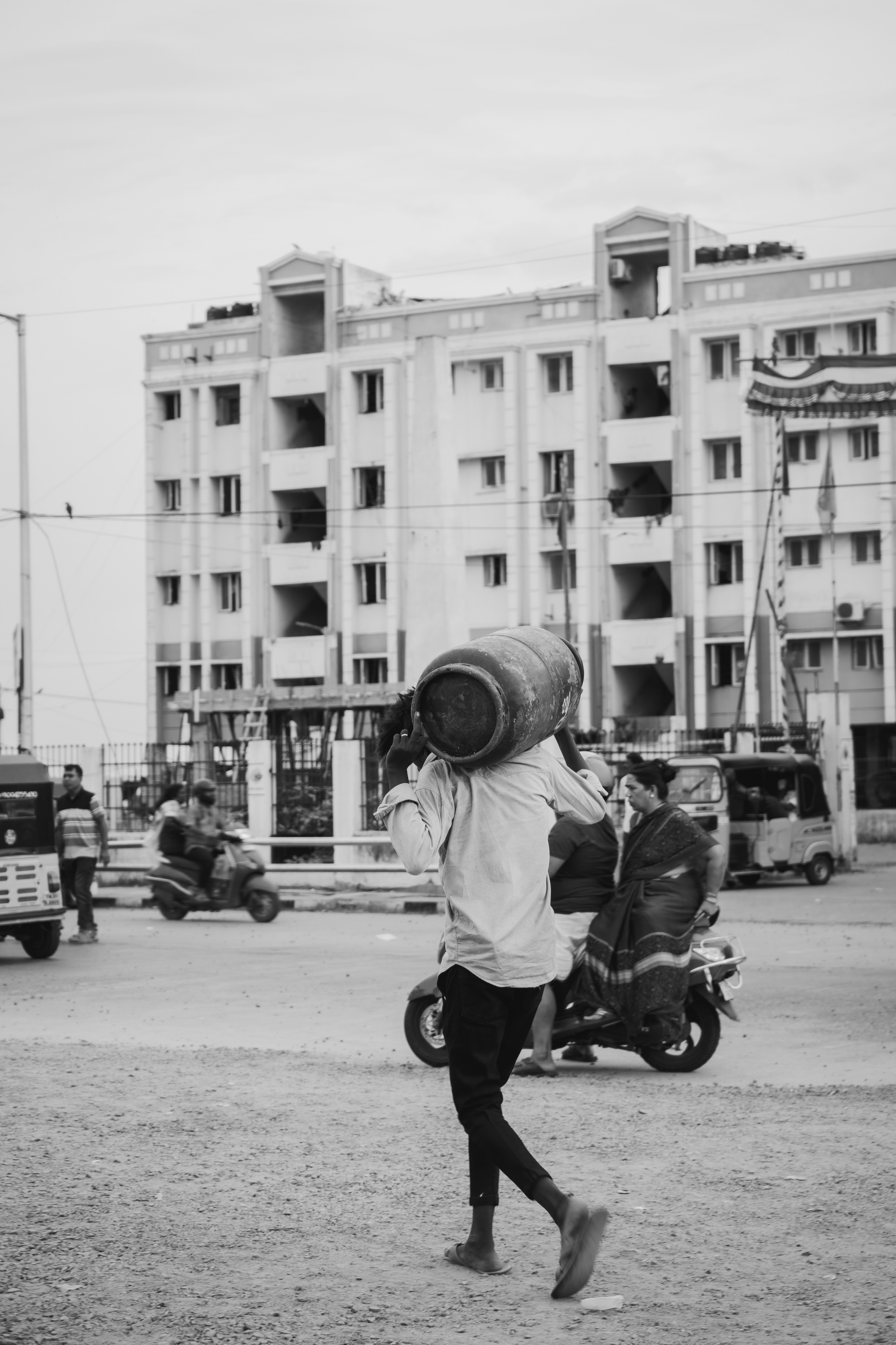 A laborer carrying a heavy gas cylinder through a bustling urban environment, showcasing the daily struggles of city life.