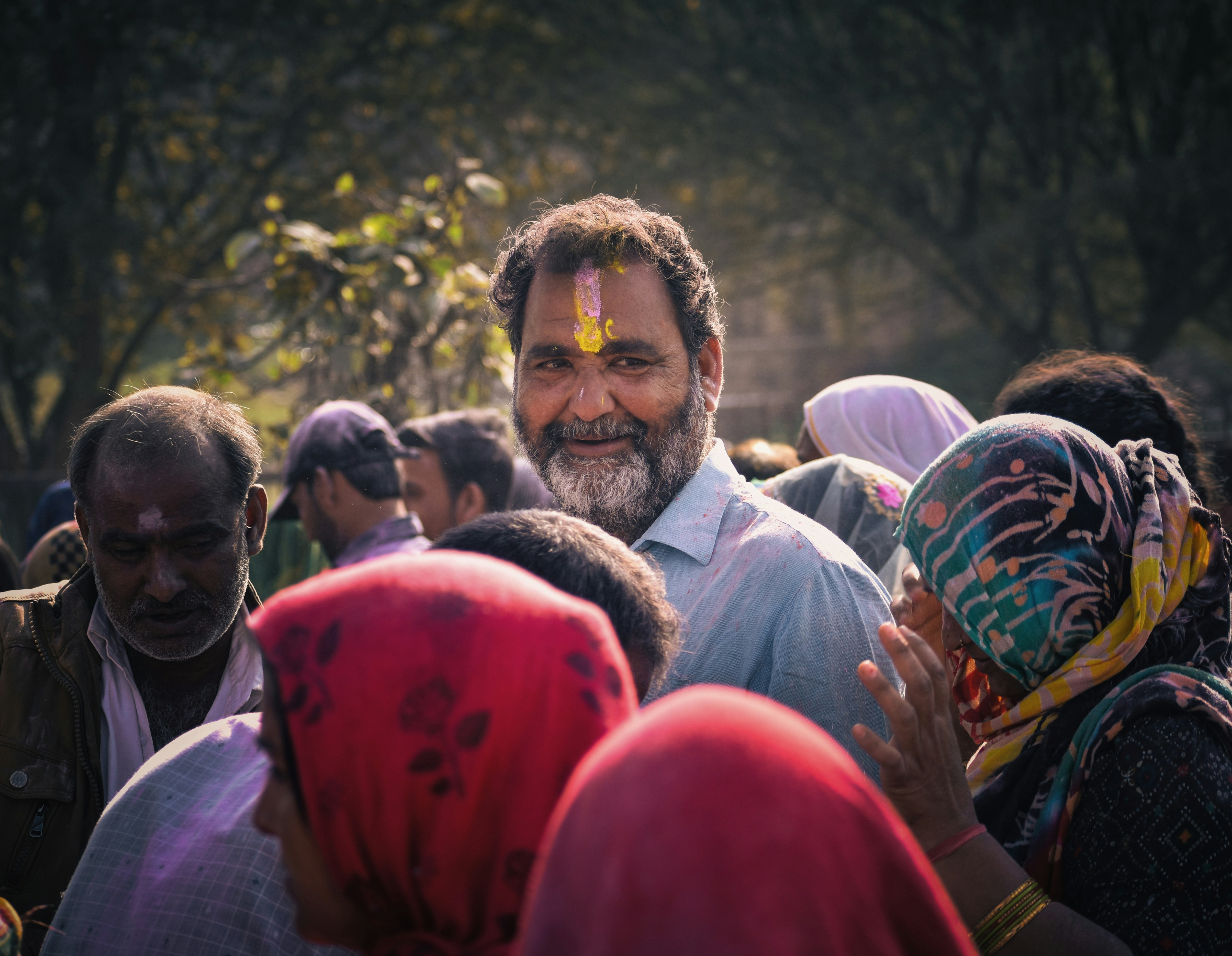 A smiling man with a vibrant yellow mark on his forehead stands amidst a crowd celebrating a festival, surrounded by colorful scarves and joyful expressions.