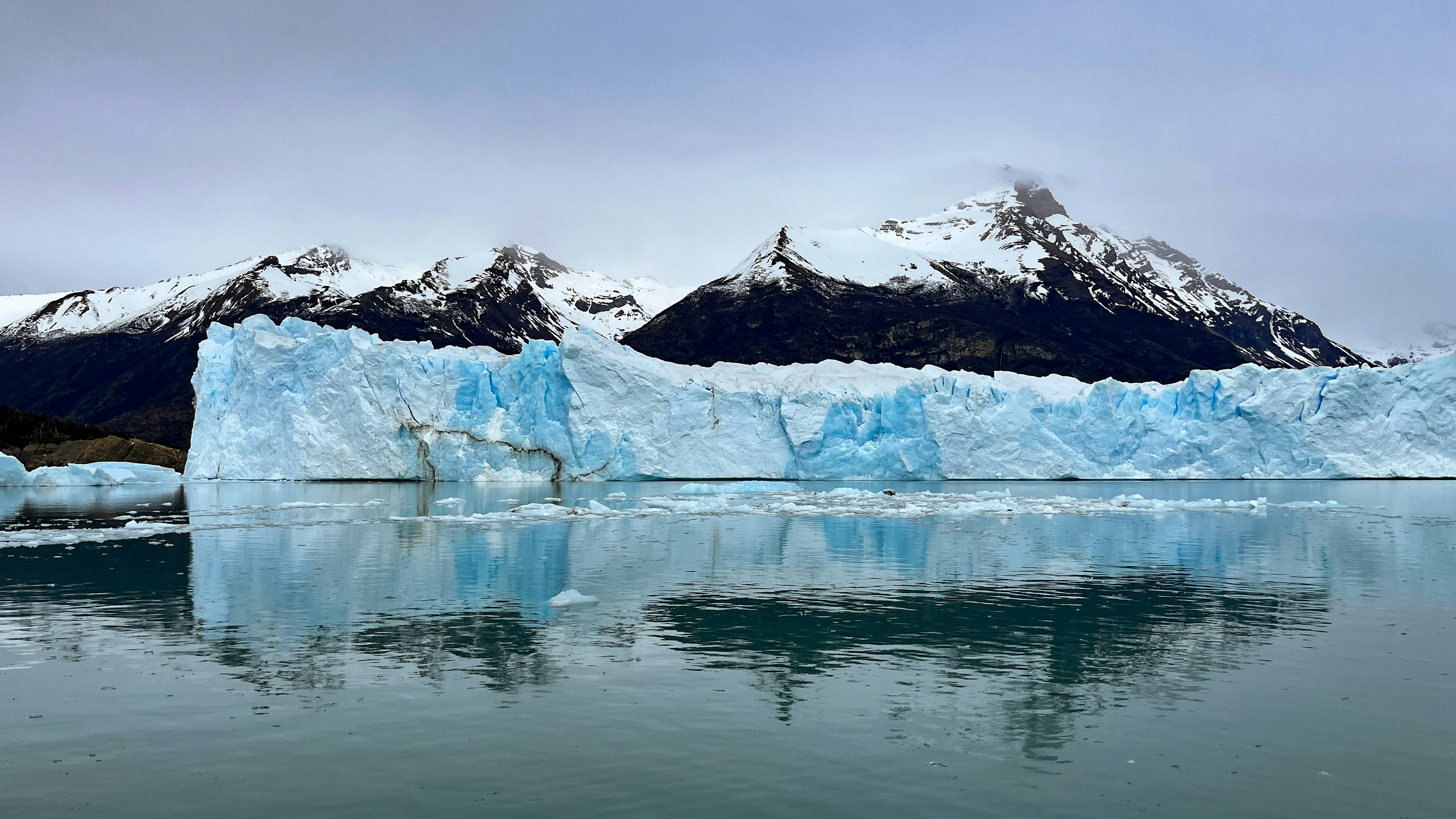 Vast glacier with rugged mountains reflected in tranquil water under a cloudy sky.