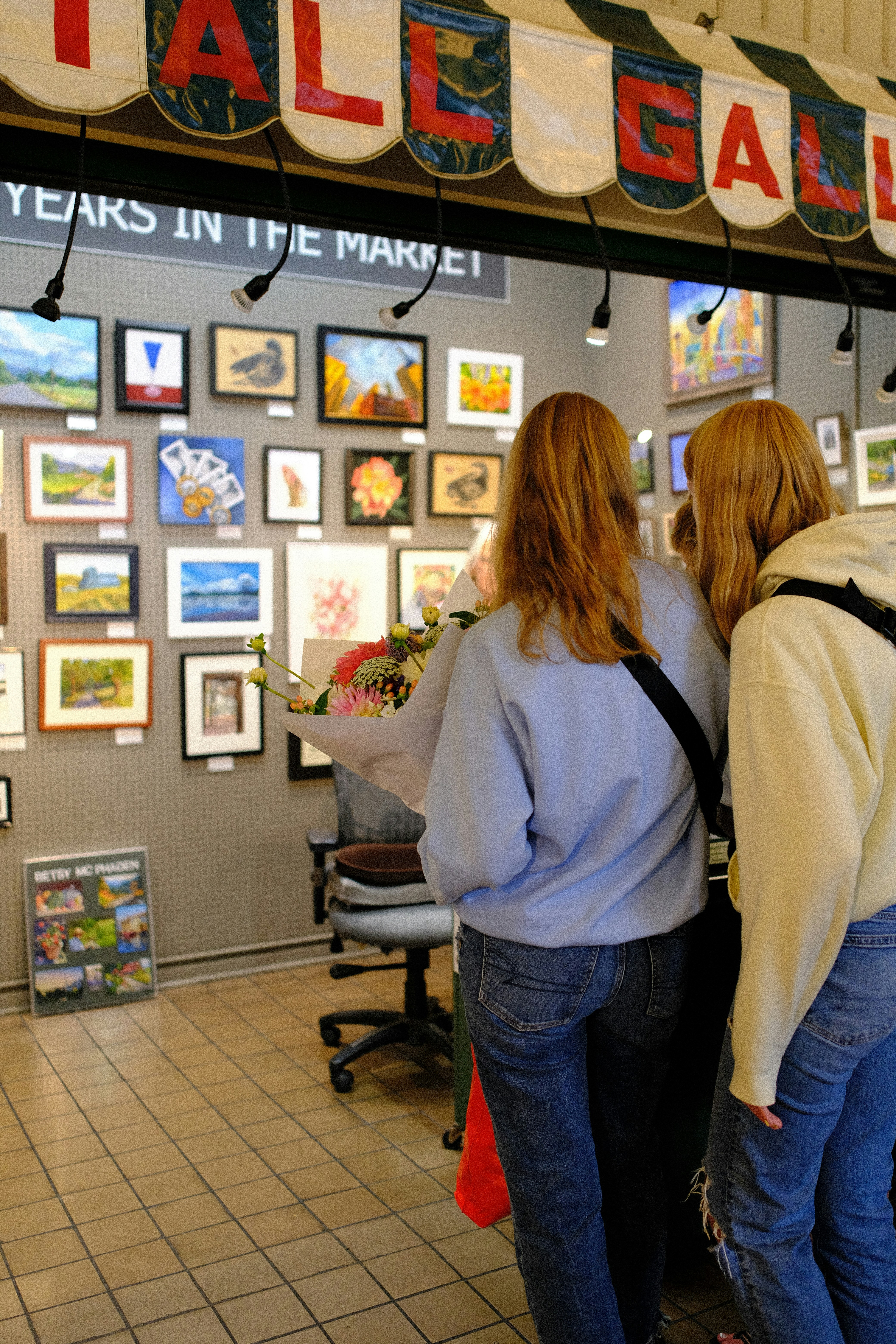 Back-to-Camera Moment: Two Women Explore a Wall of Framed Art in a Bright Gallery