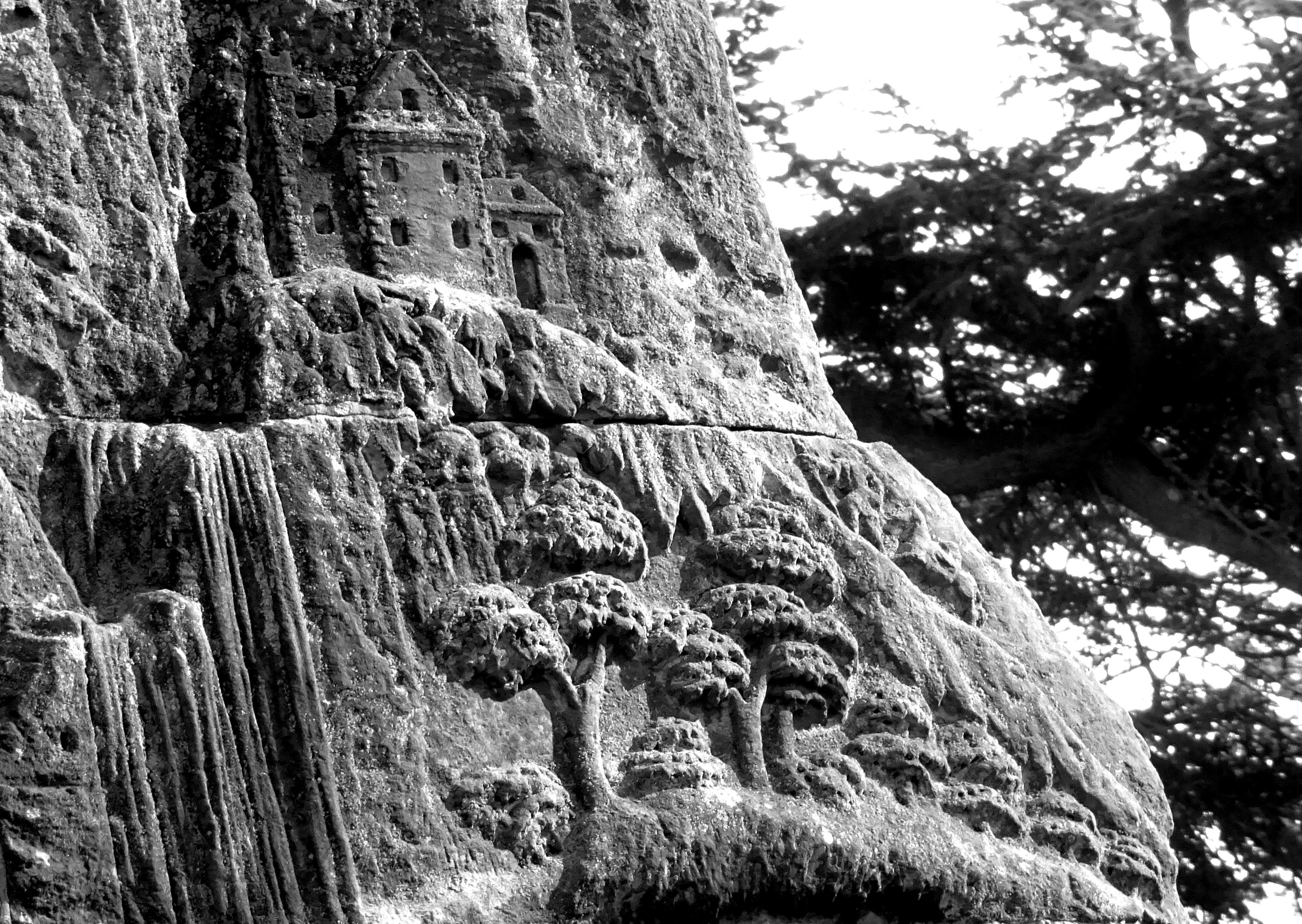 Intricate stone carvings depicting a house and trees, showcasing artistry on a rock face. The monochrome palette enhances the details of the sculpture.