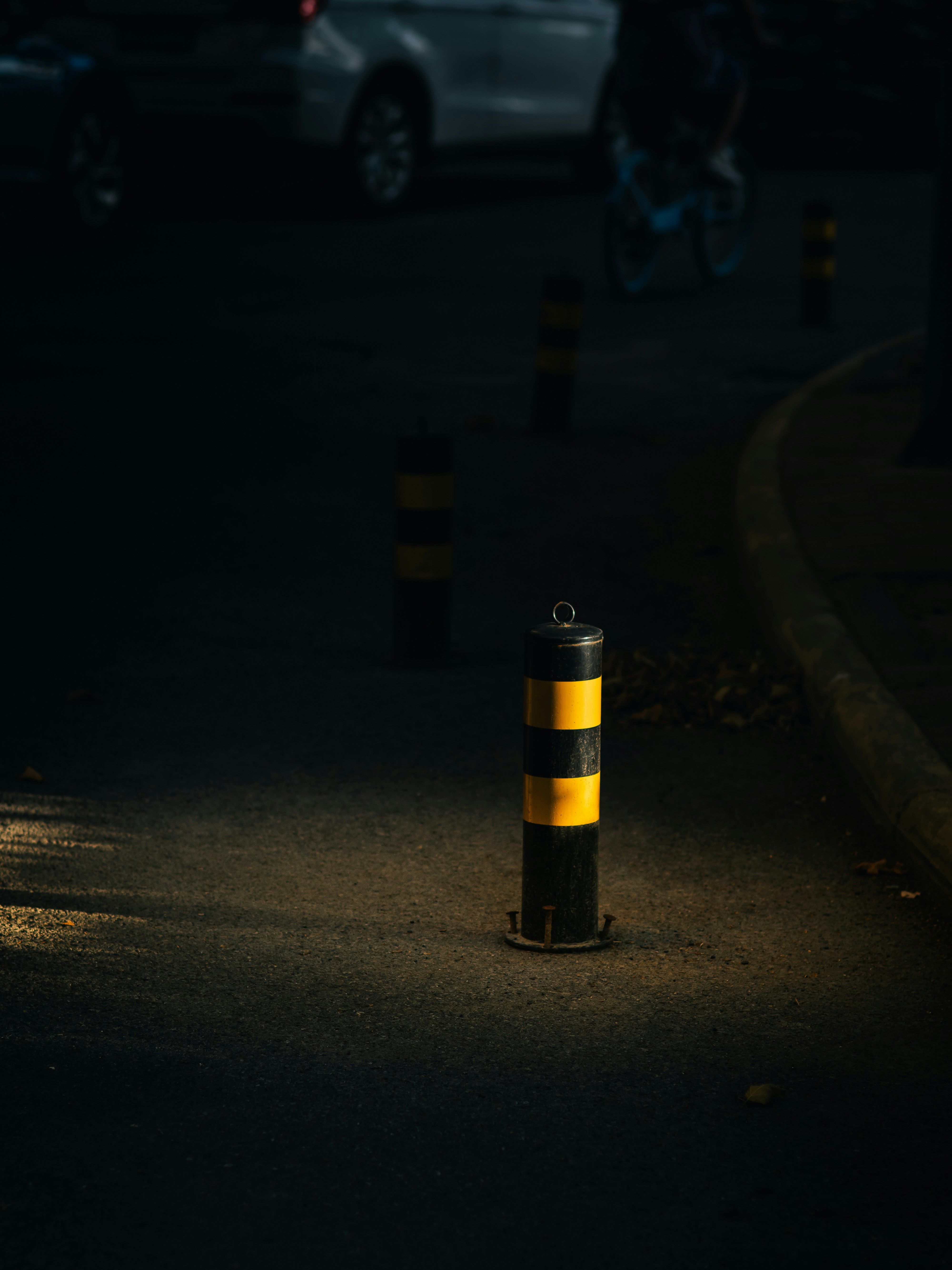 Night-time street photograph focusing on a yellow-and-black striped bollard bathed in a narrow beam, with a blurred car and cyclist in the background.