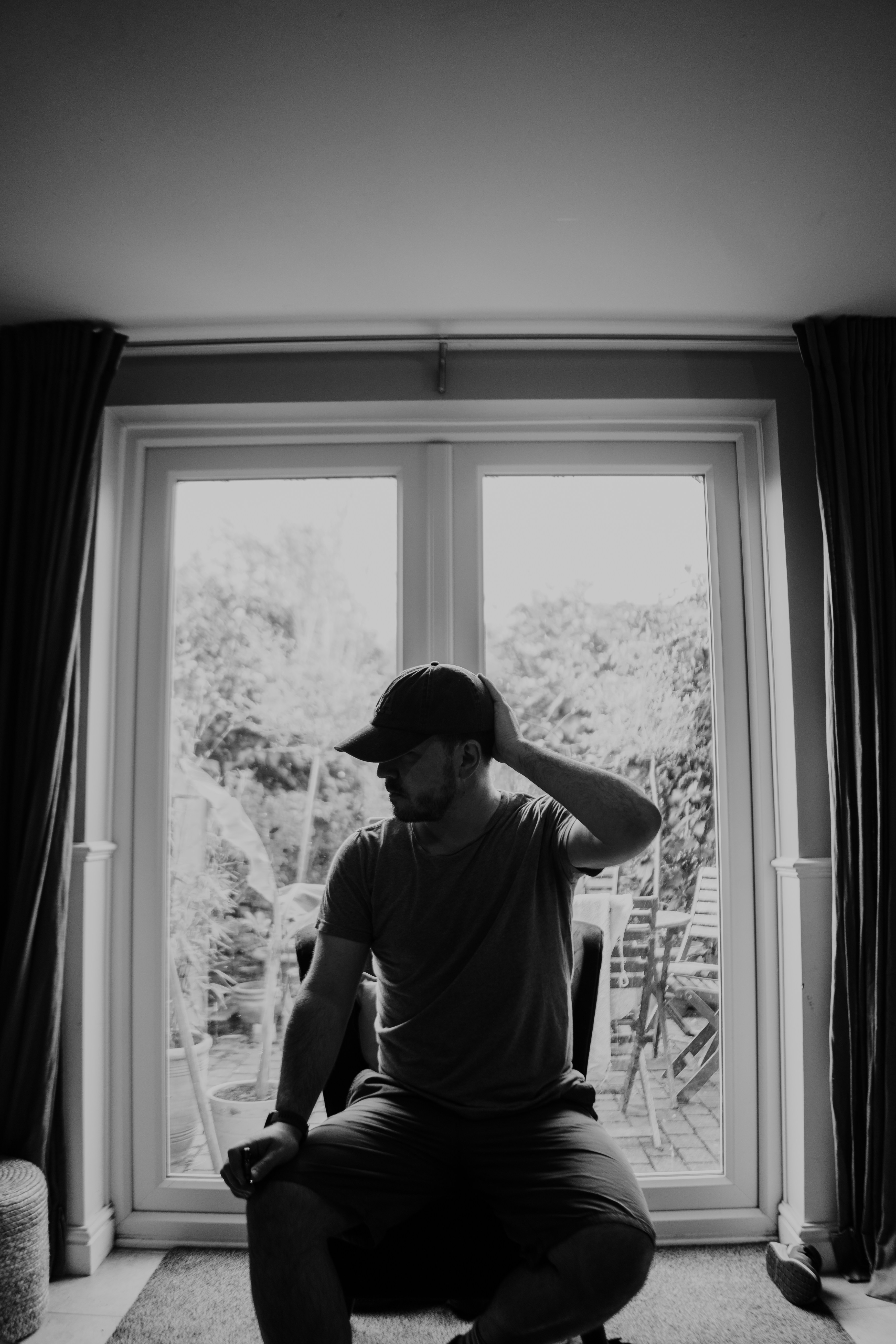 Person sitting in front of a large window, softly illuminated by natural light, creating a moody contrast with the bright garden outside.