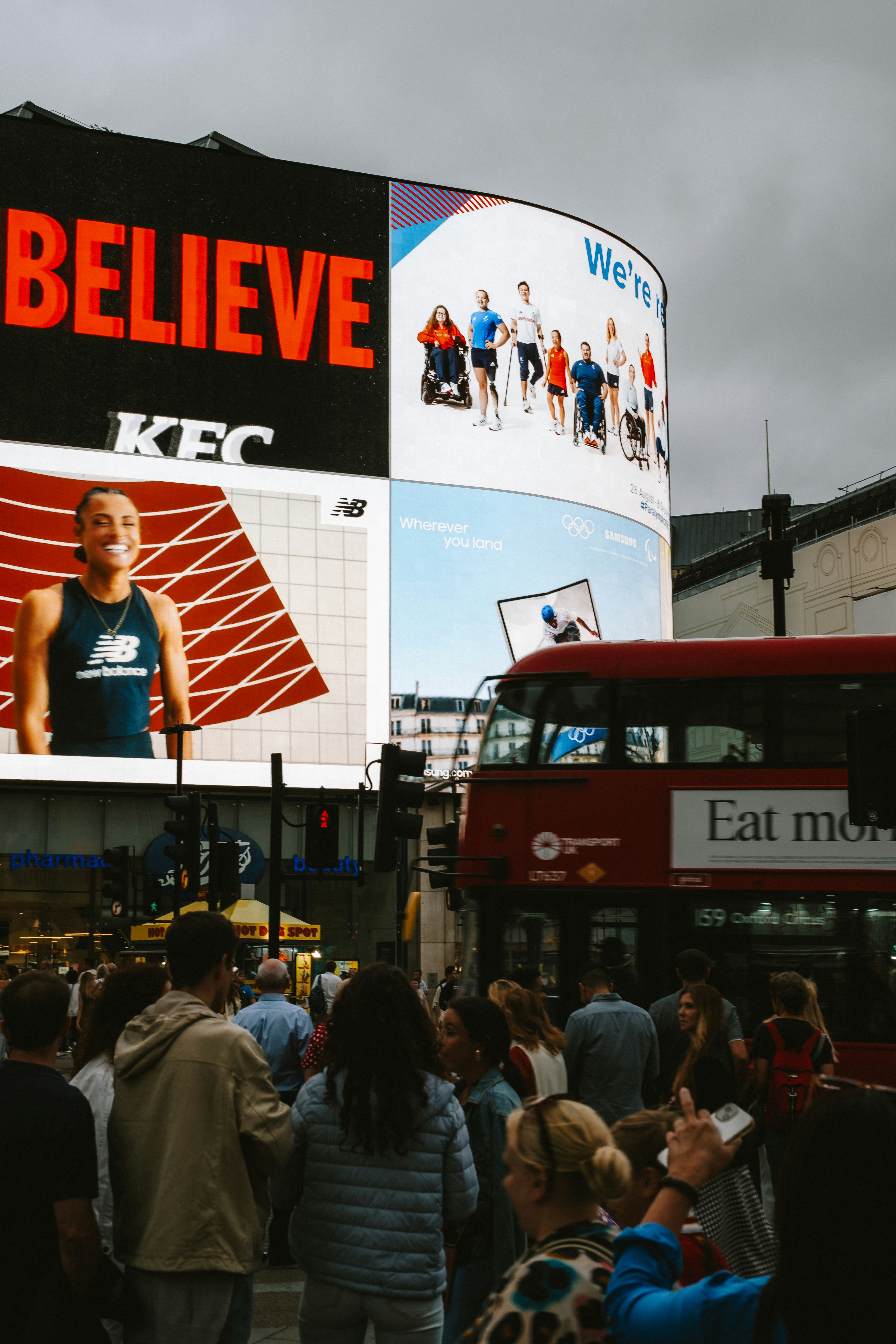 Crowd of pedestrians navigating a busy city intersection, with large digital billboards displaying advertisements above. The scene captures the dynamic energy of urban life.