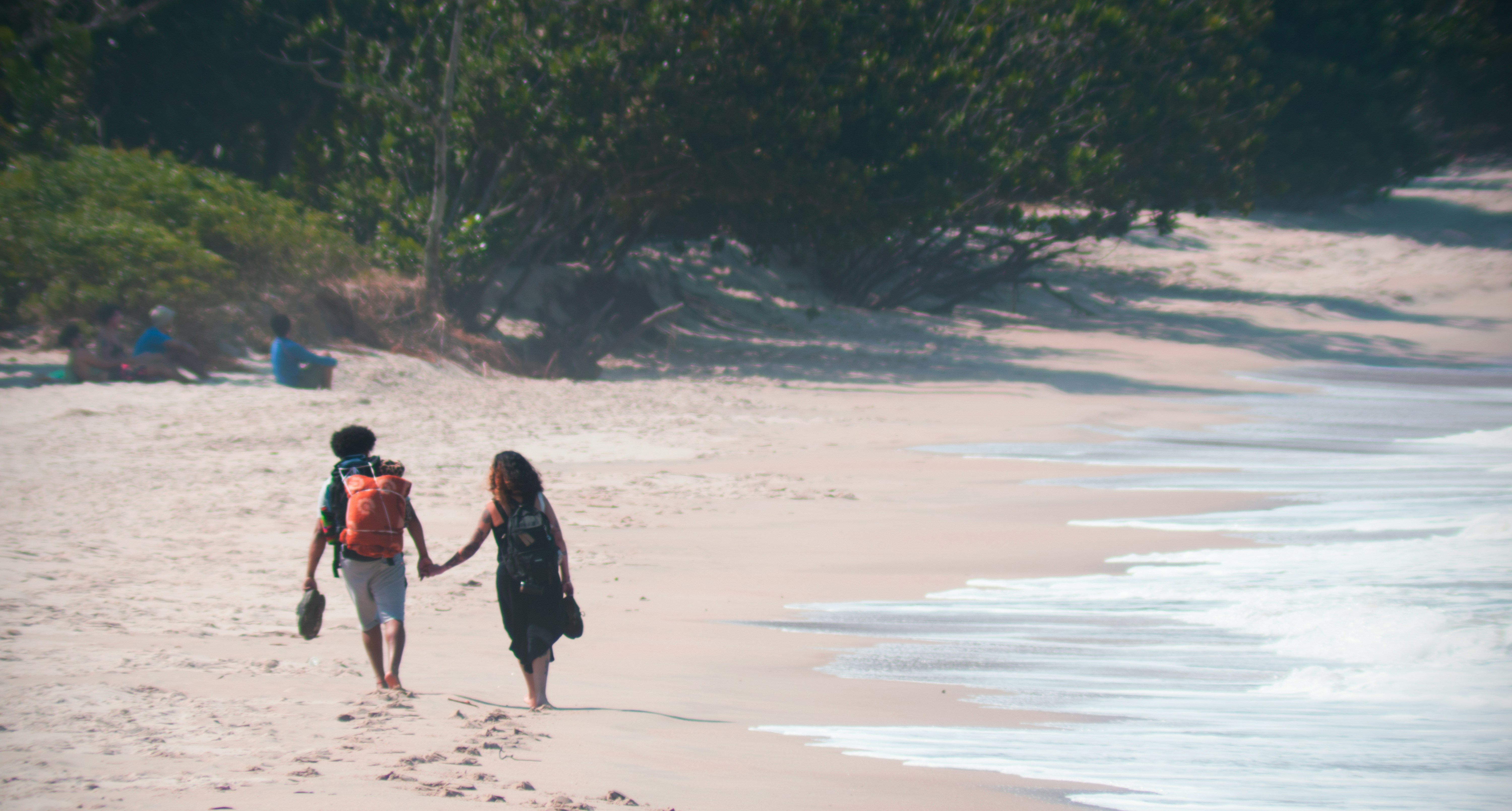 Two people walk hand in hand along a sunlit beach with gentle waves lapping at the sand.