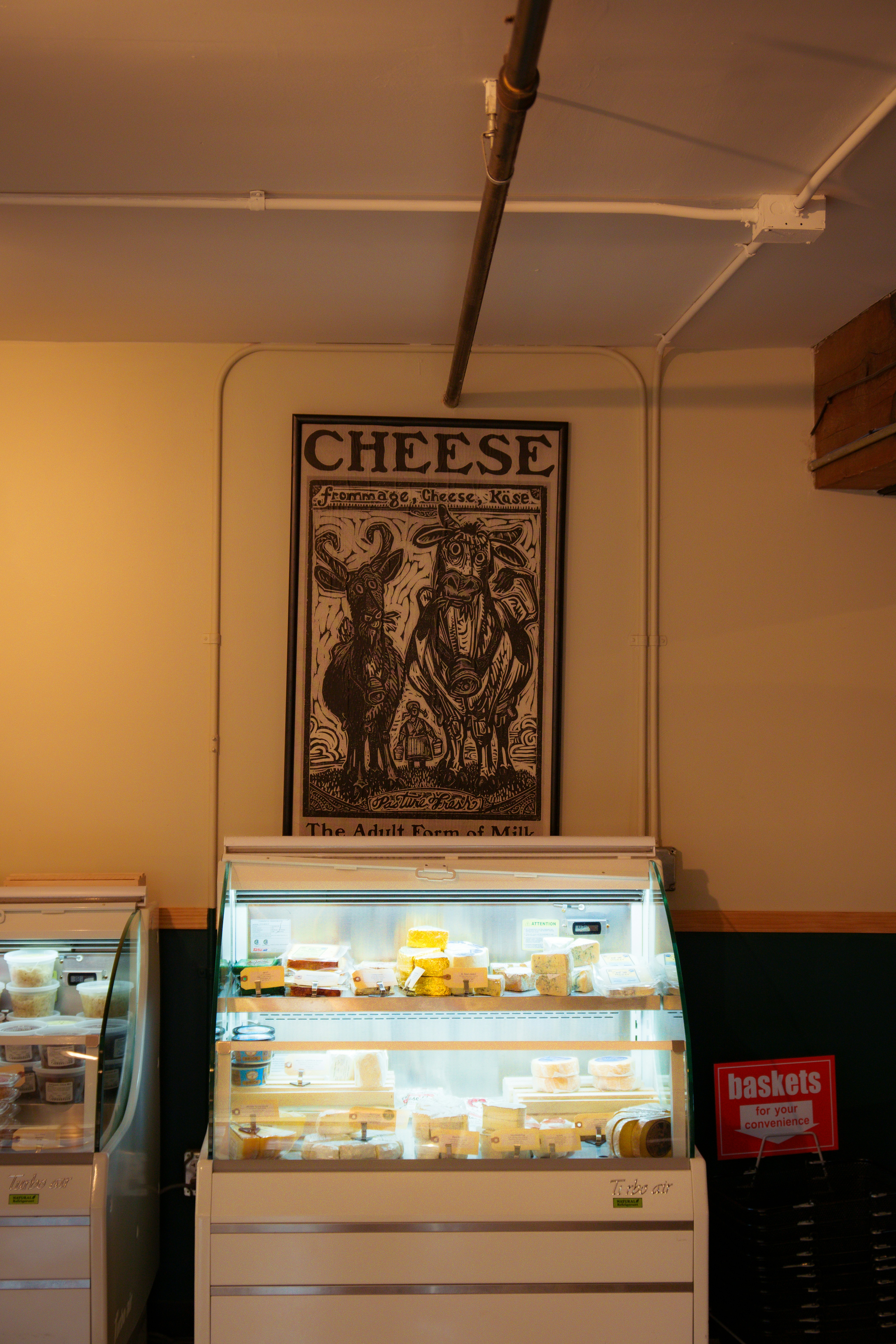 A softly lit cheese display case sits under a framed vintage poster in a small shop, with shelves of cheese and dairy goods.