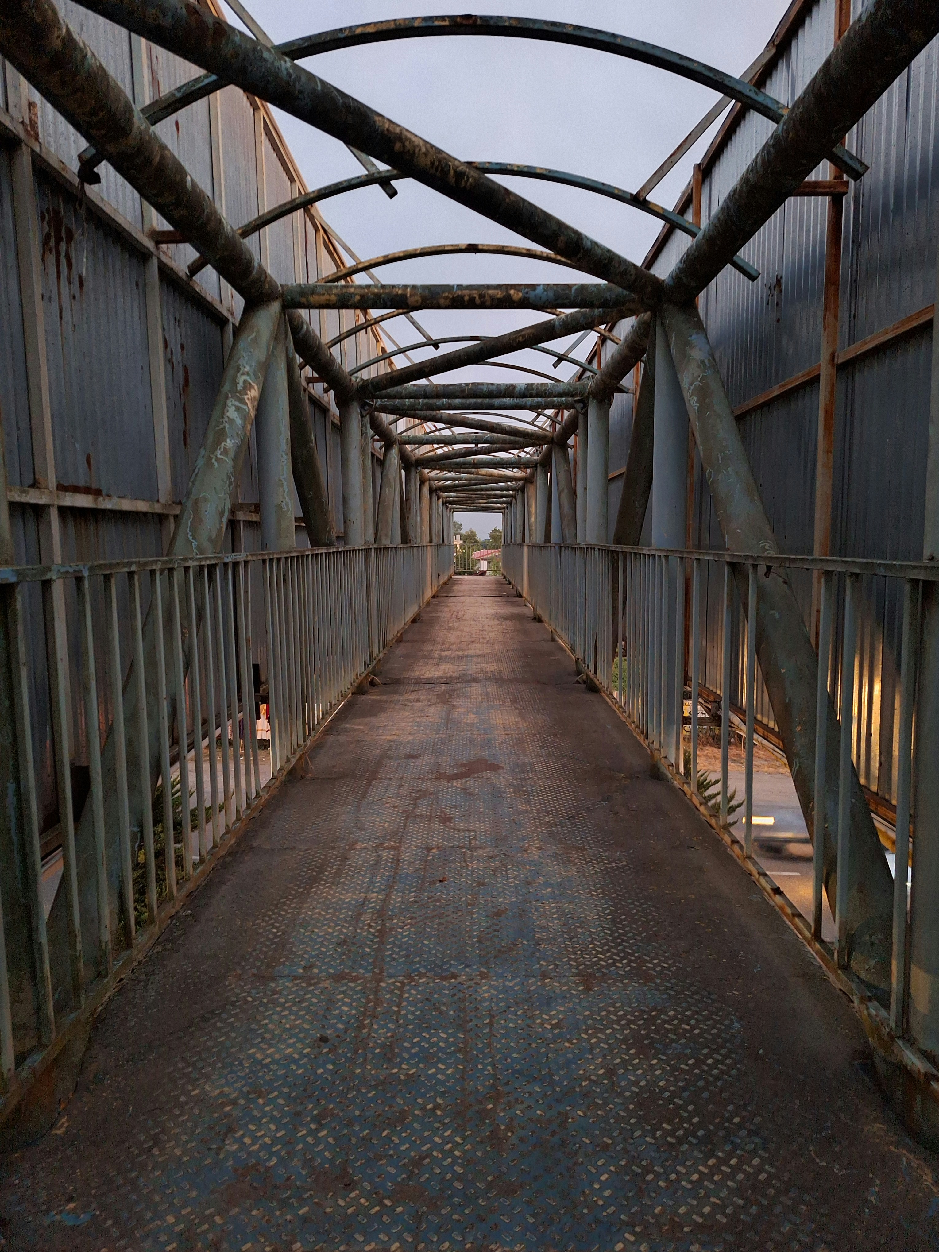 Weathered metal pedestrian bridge with arched overhead ribs creates a strong vanishing-point perspective toward a distant glow at the end.