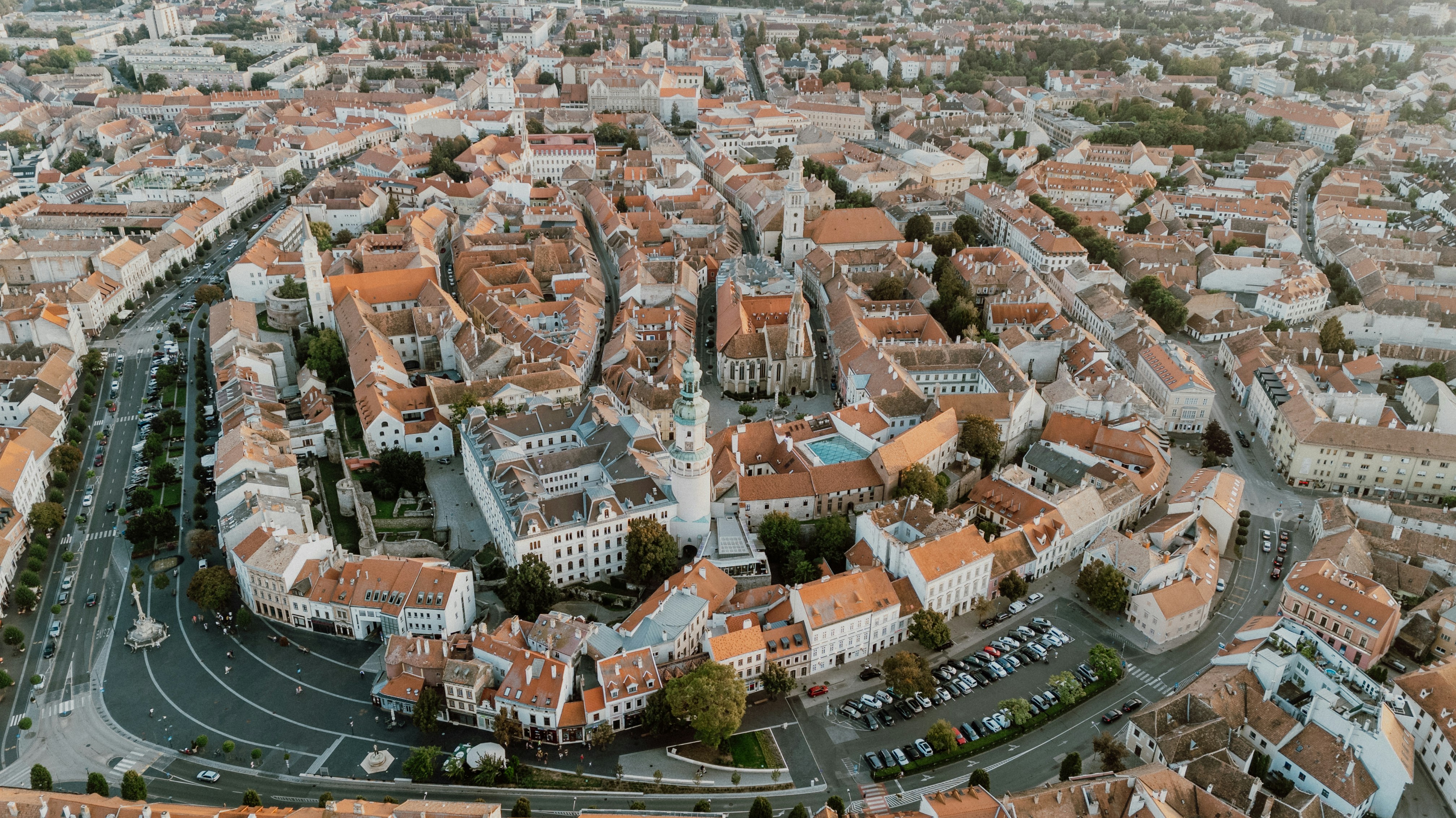 Aerial view of a European town with densely packed red-tiled roofs, a circular plaza, and a white church spire at the center. Streets radiate from the plaza, weaving intricate patterns across the urban fabric.