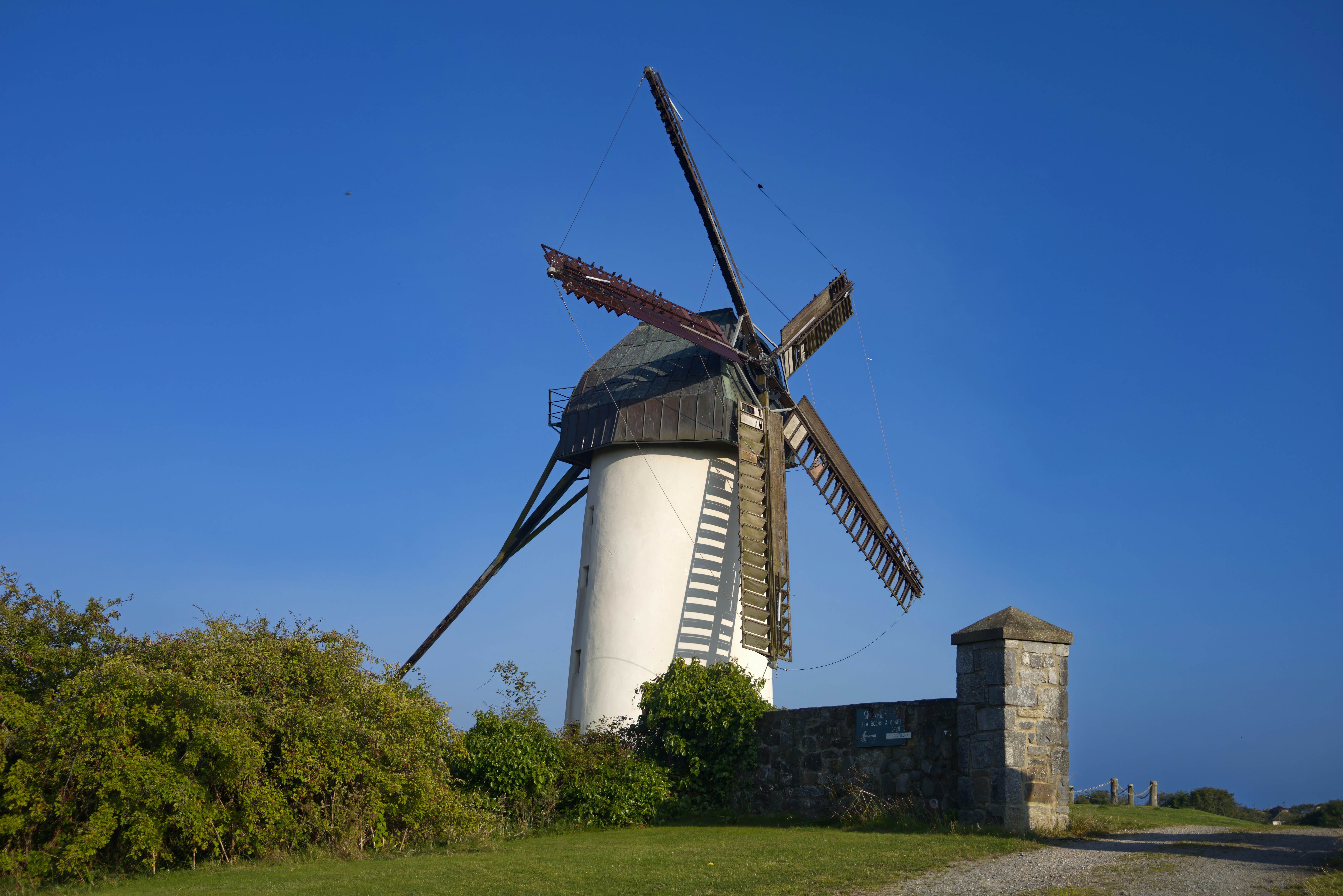 Traditional windmill with large sails stands beside a stone wall under a clear blue sky.