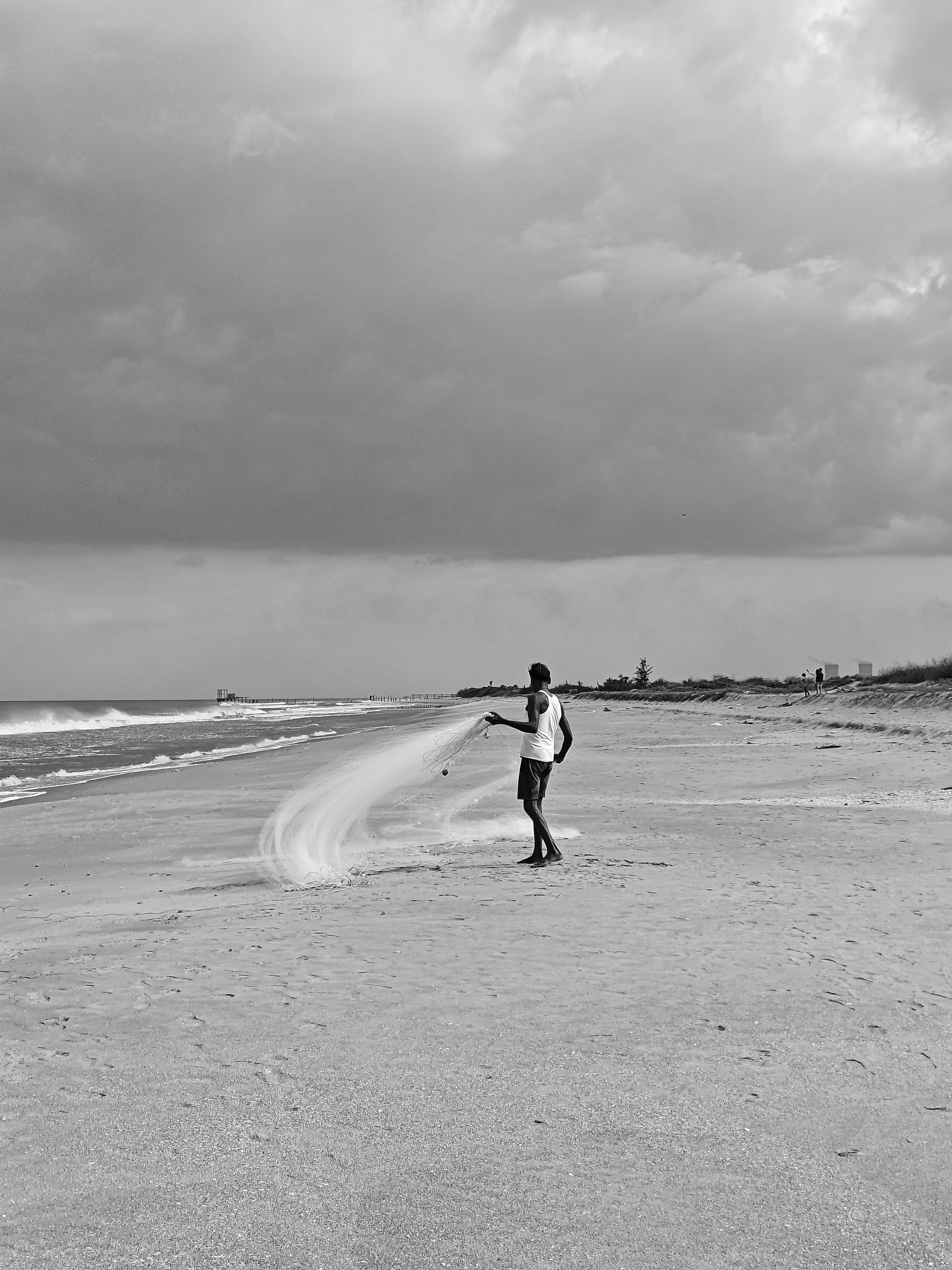 Black-and-white photograph of a lone figure on a quiet beach releasing a spray that curves through the air against a moody, overcast sky.