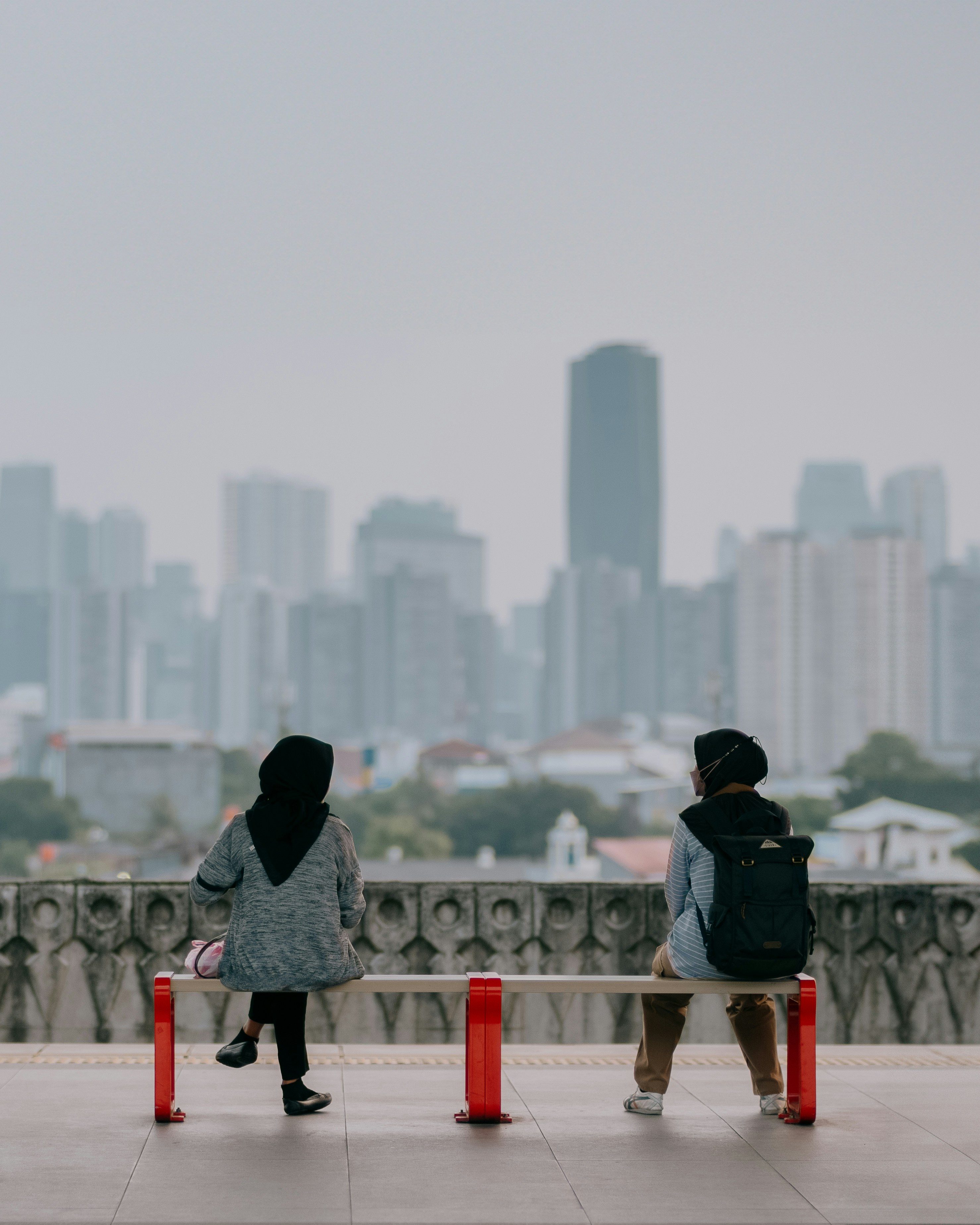 A touching moment between a mother and her child, both wearing headscarves, sitting on a bench at Manggarai Station, Jakarta. They face away from the camera, gazing at the distant Jakarta skyline. The scene exudes a sense of calm and contemplation, with the urban landscape providing a stark contrast to their quiet moment.