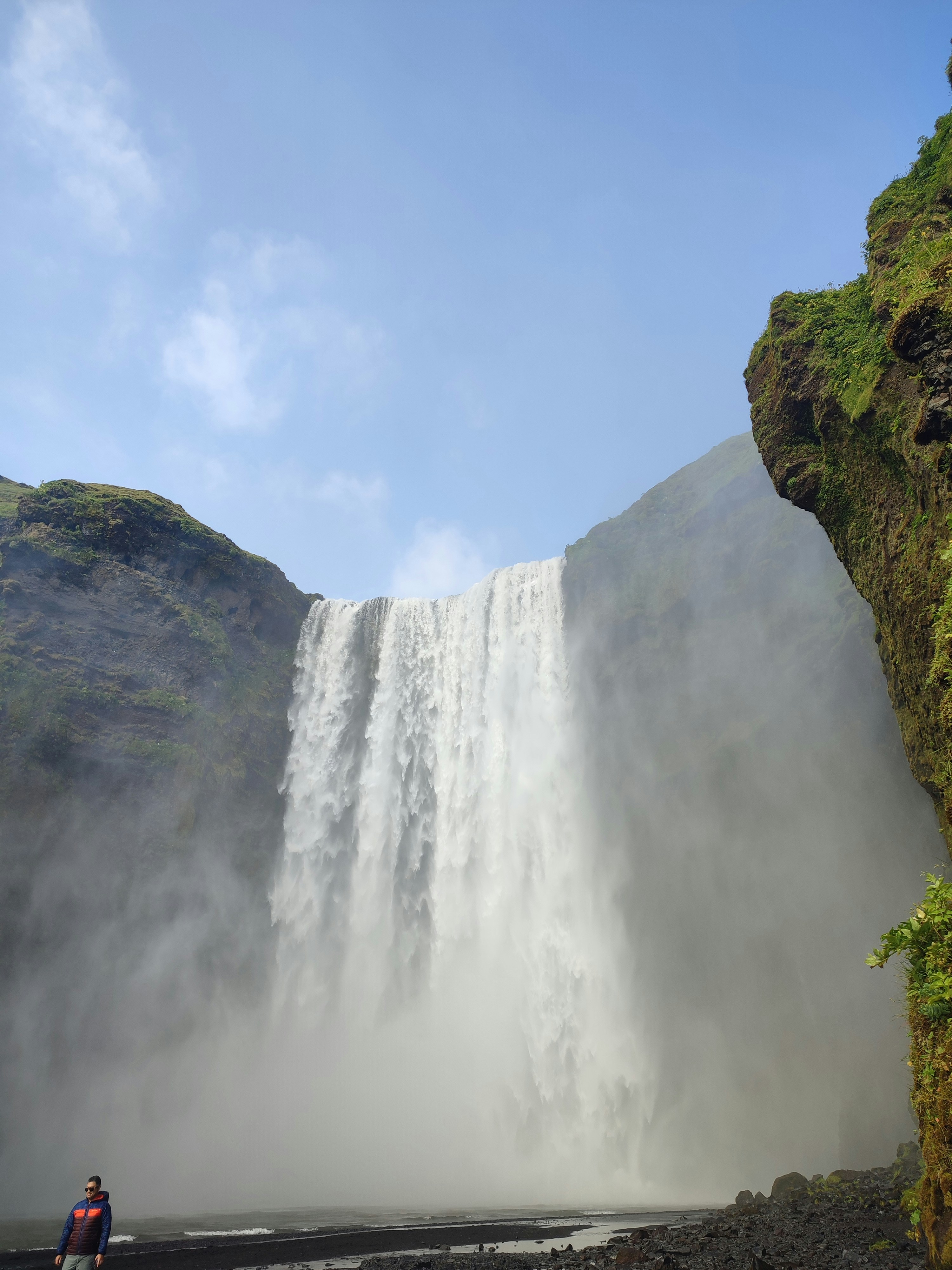 Tall waterfall cascades between mossy cliffs as mist swirls at the base. A lone observer on the shore provides scale.