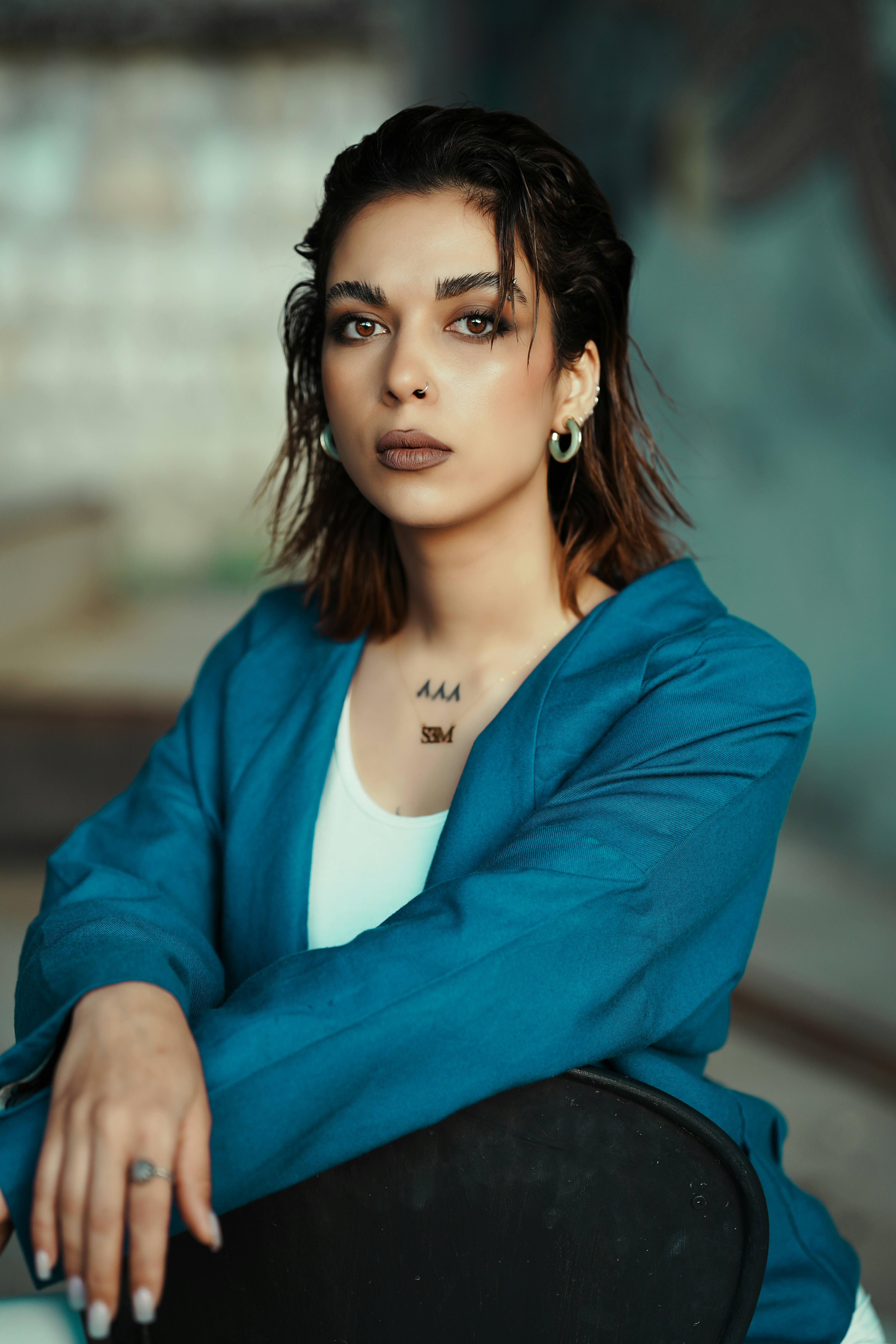 A young woman in a striking blue blazer sits confidently on a chair, showcasing her unique style and tattoos against a softly blurred background.