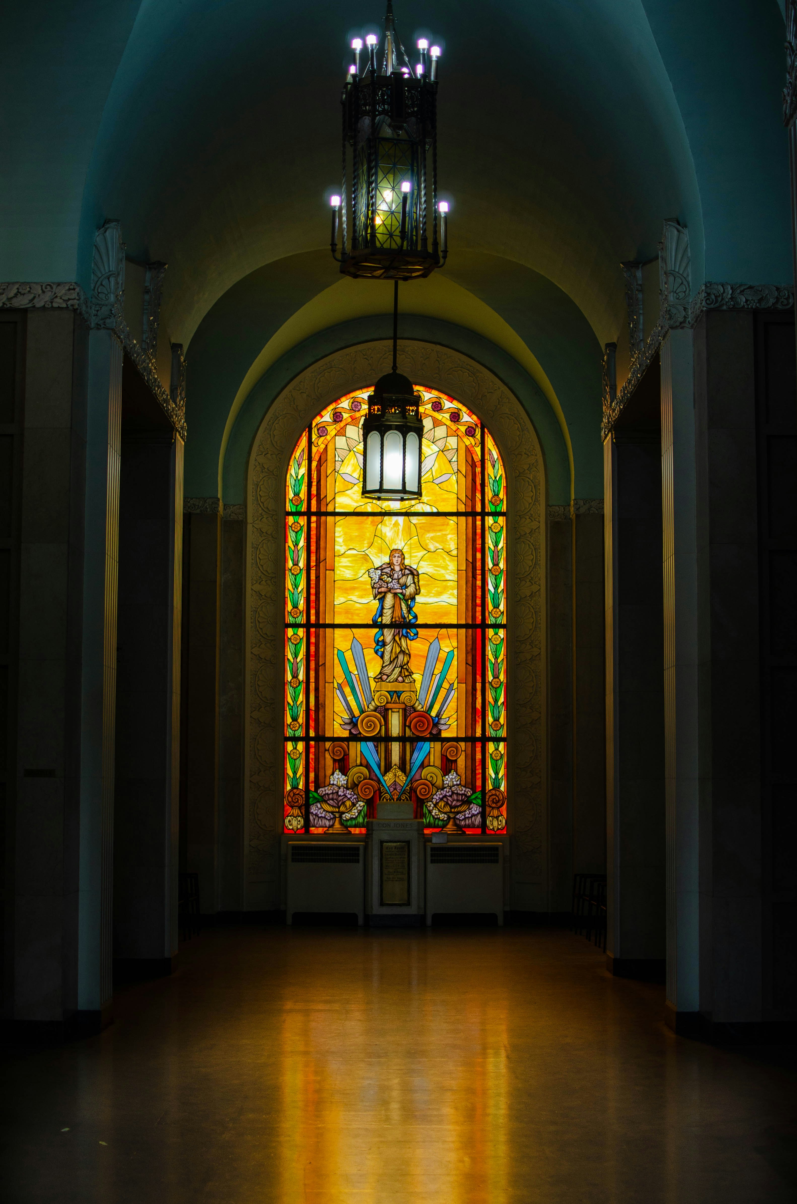 An exquisite stained glass window in a historic mausoleum, featuring vibrant colors and intricate religious artwork. The warm glow of the window reflects on the polished floor, creating a serene and reverent atmosphere. Ideal for stock photography buyers looking for images of religious art, church interiors, and historic architecture. Perfect for use in publications, religious-themed projects, or as a visual element in design work focusing on spirituality and heritage.