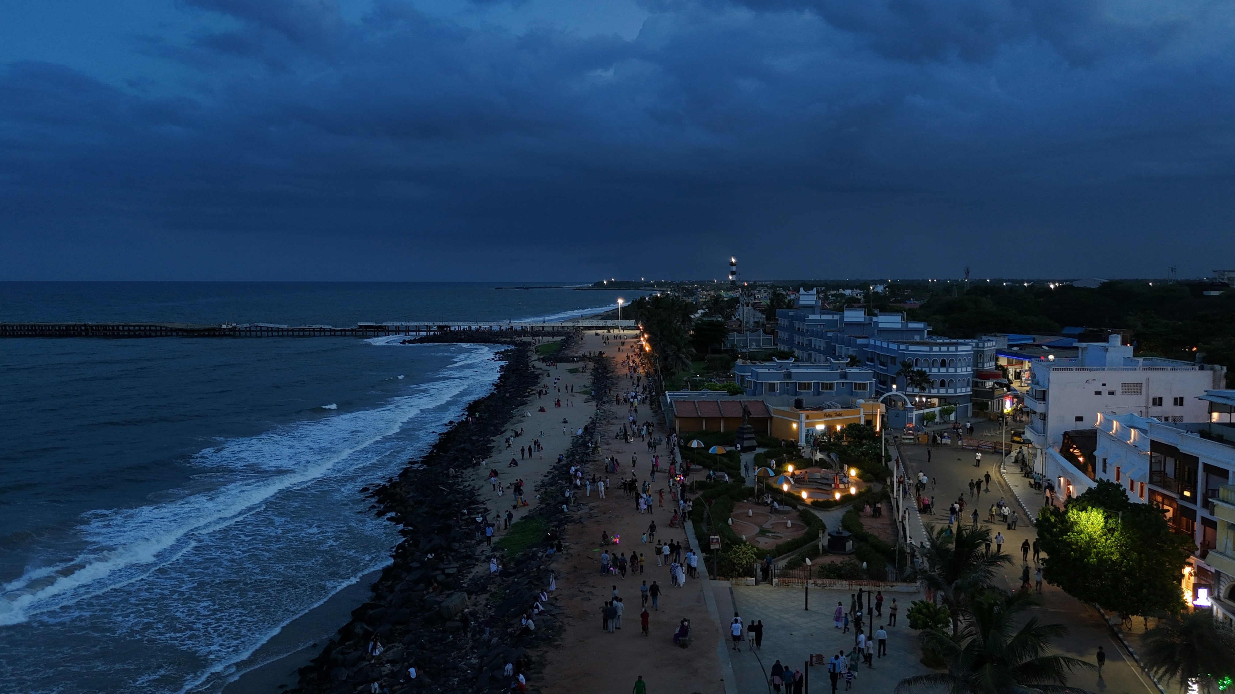 Coastal promenade bustling with evening strollers, illuminated by warm lights against a backdrop of a darkening sky and gentle waves.
