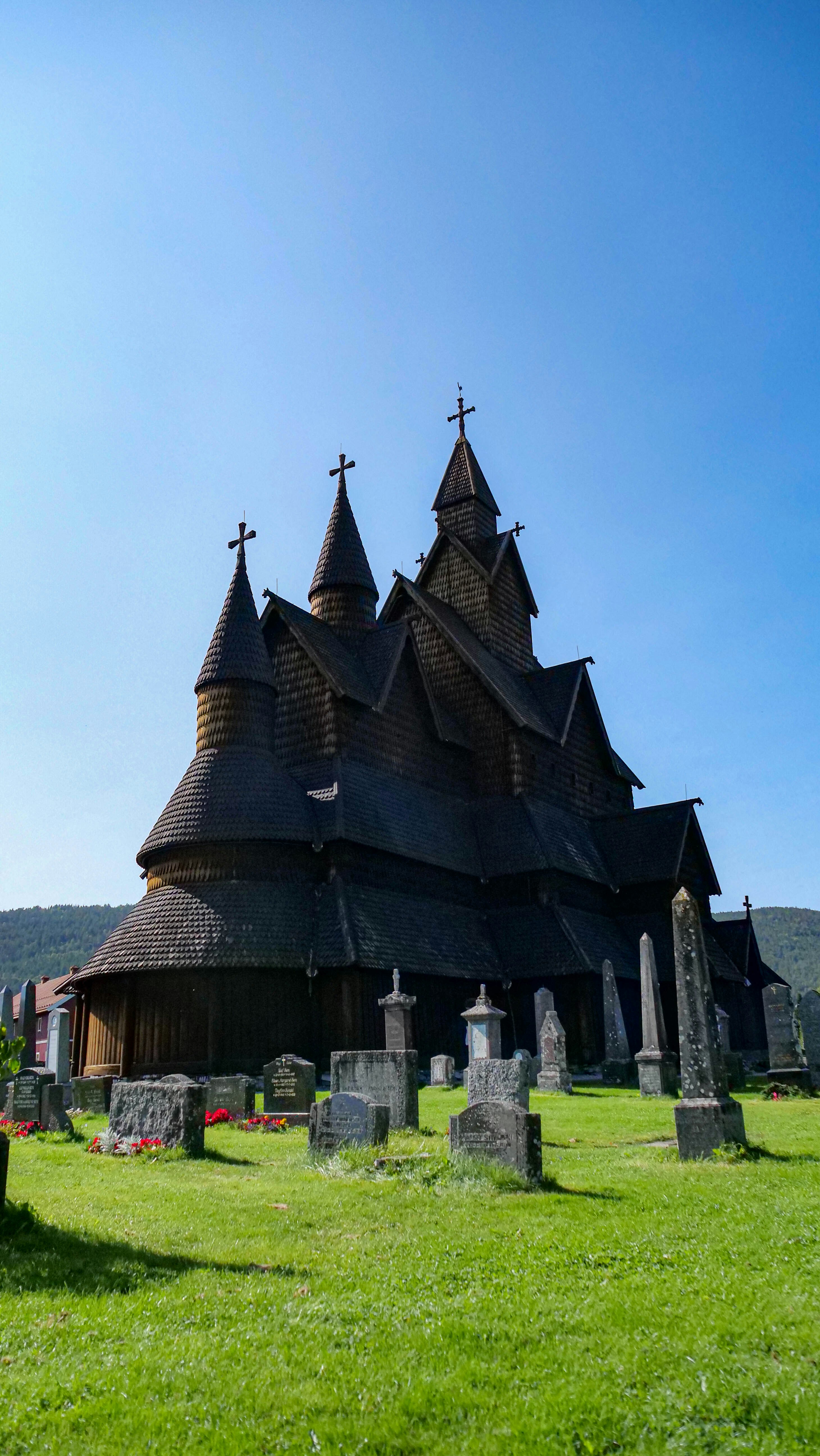 A majestic stave church with multiple spires stands proudly among gravestones, surrounded by lush green grass and a clear blue sky.