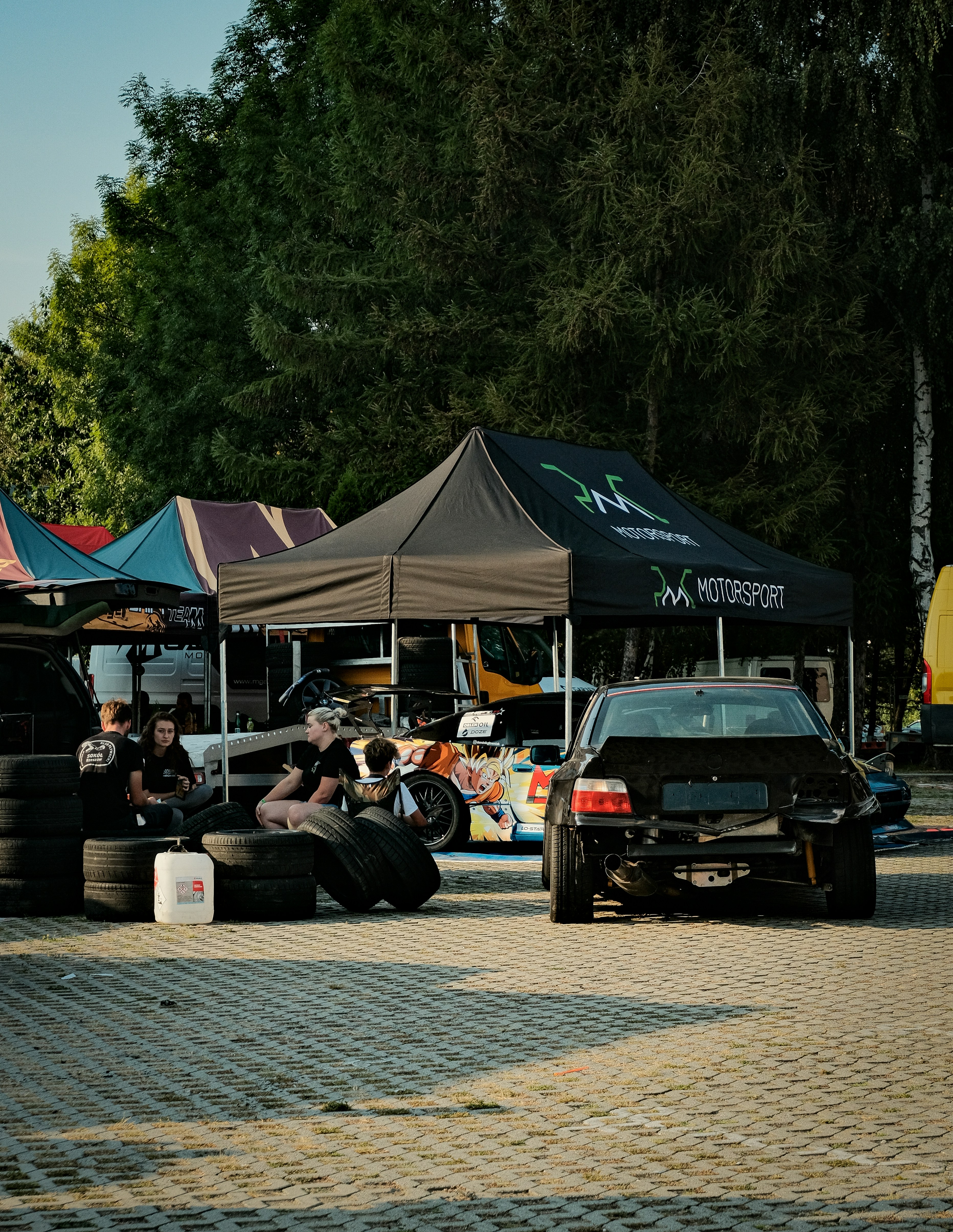 Team tents and a black car sit in a paddock with spare tires and a colorful race car under a canopy. The scene conveys the pit area atmosphere of a motorsport event.