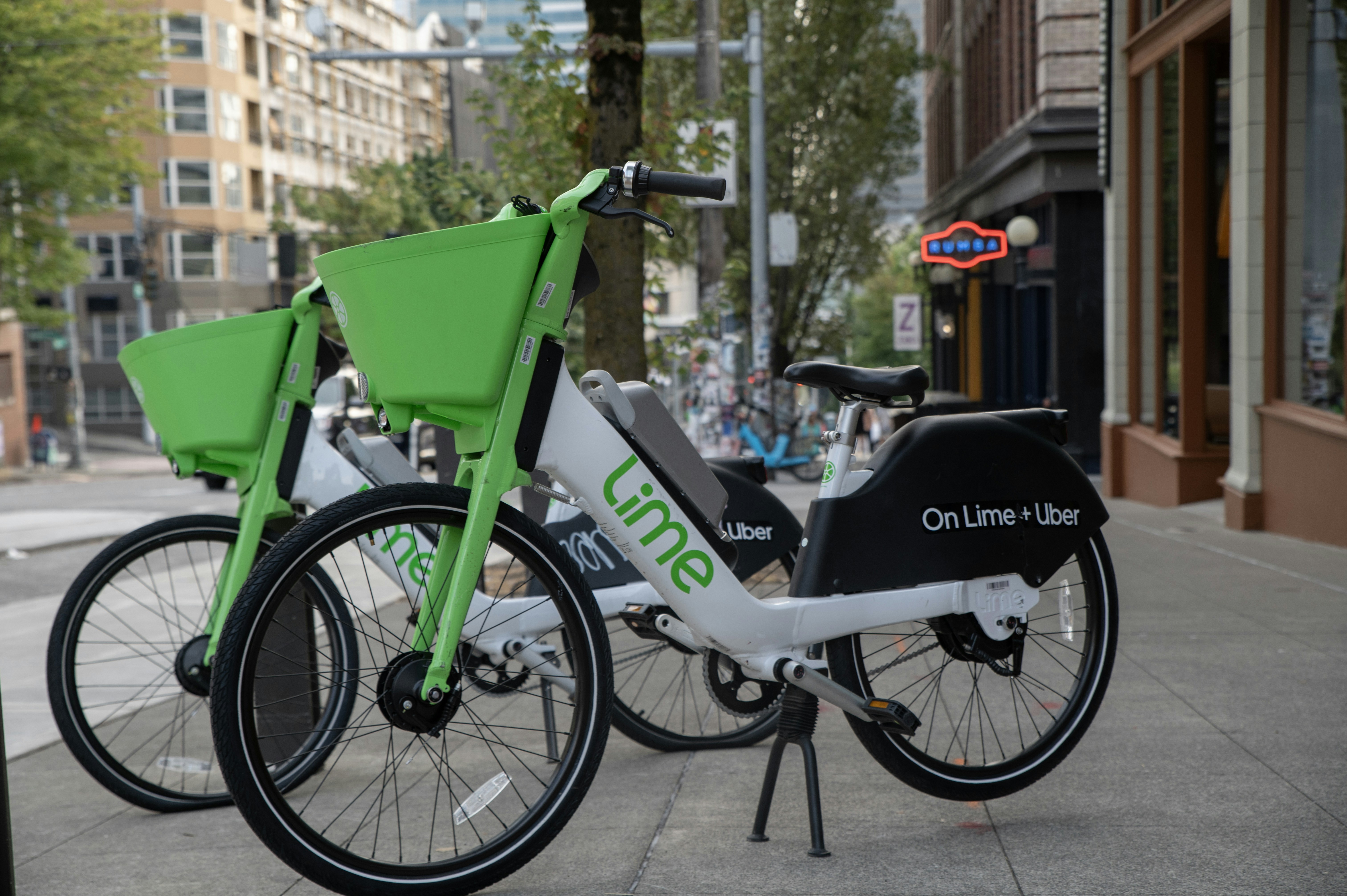 Two Lime bicycles parked on a city sidewalk, showcasing a shared-mobility setup with branded bikes and a blurred urban background.