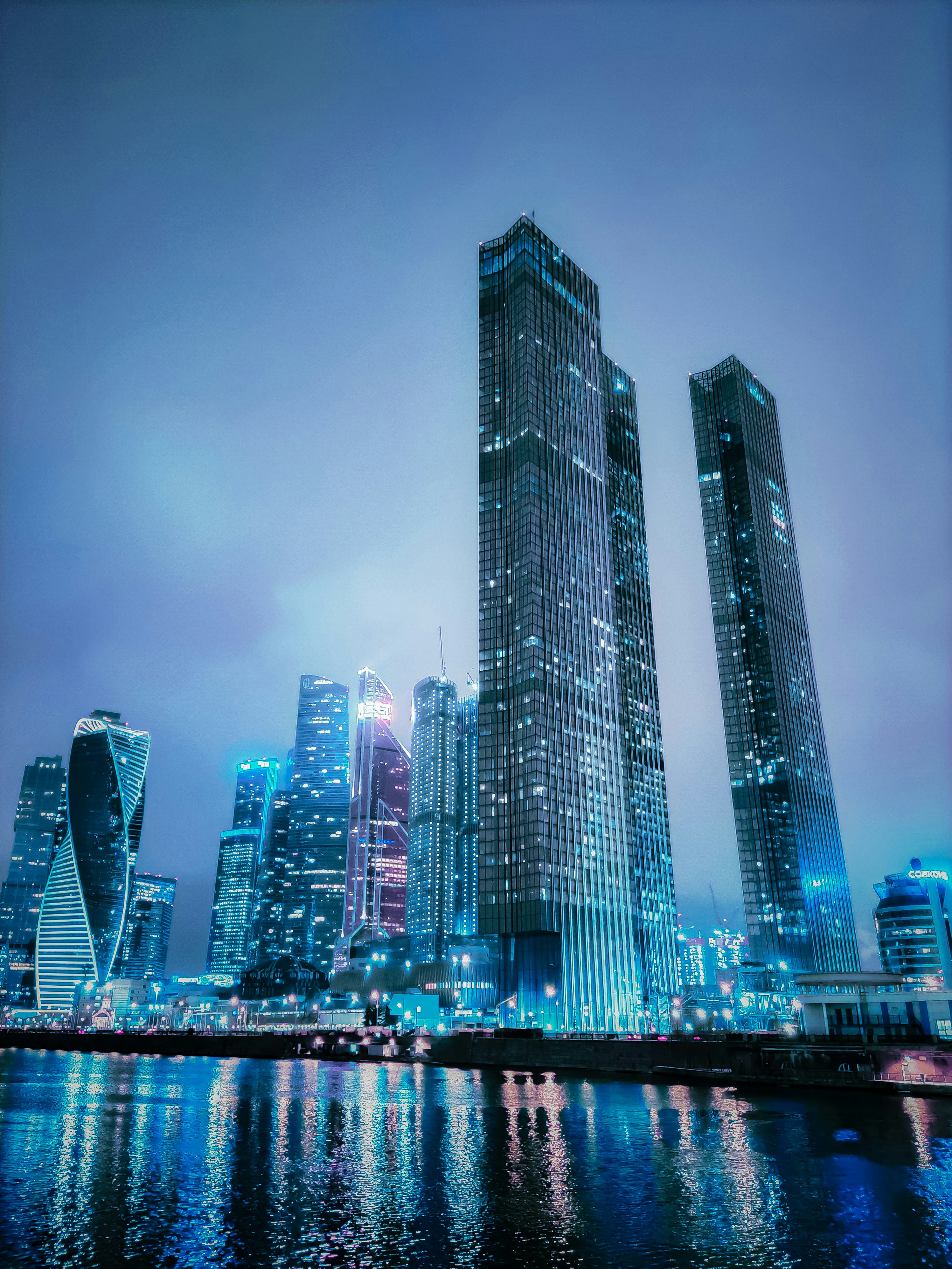Night cityscape photograph of futuristic skyscrapers reflecting on a calm river, bathed in cool blue and cyan lights.