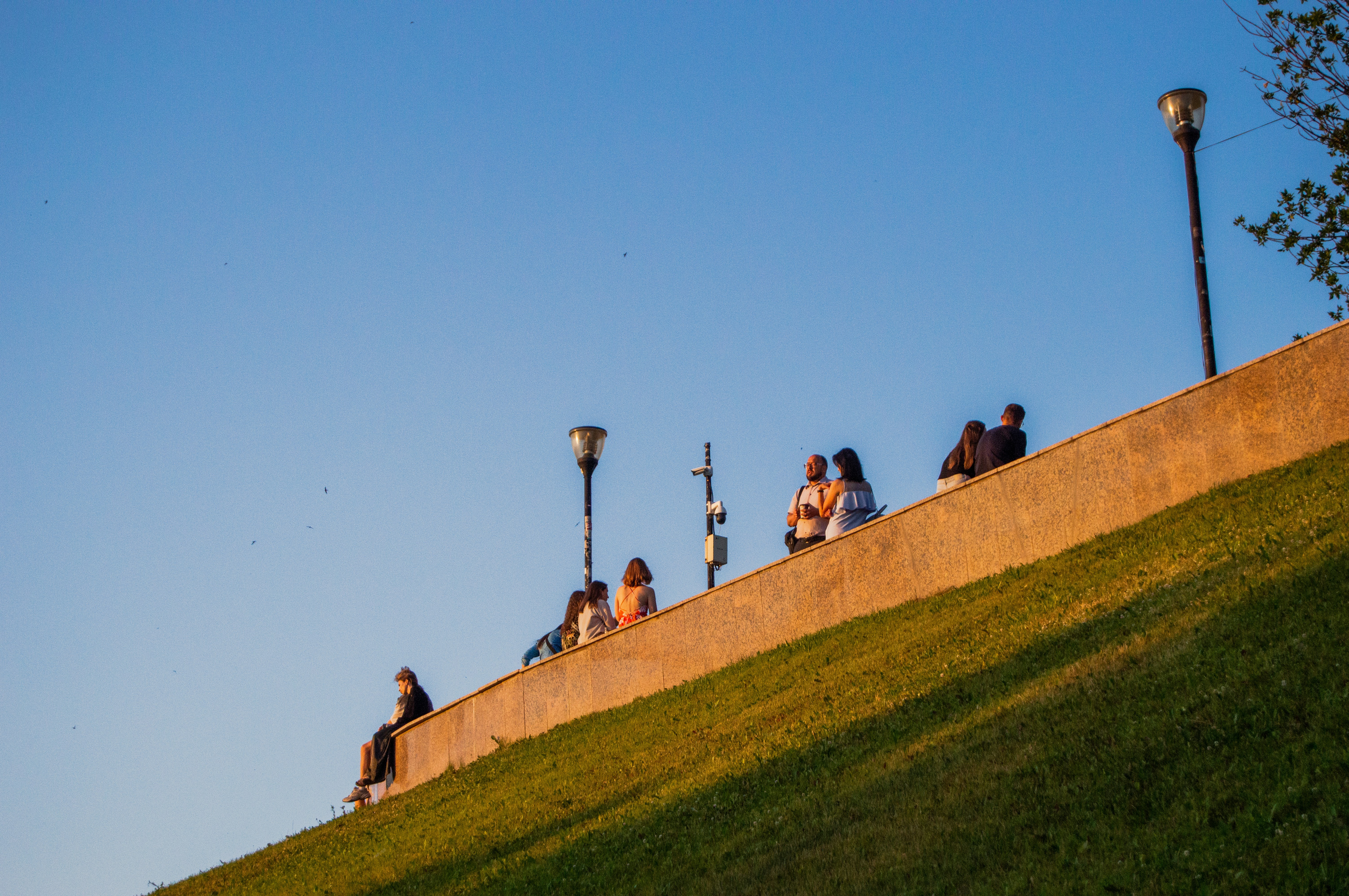 A group of people enjoying the sunset on a grassy slope, with lampposts illuminating the scene. The warm glow of the evening light adds a serene atmosphere.
