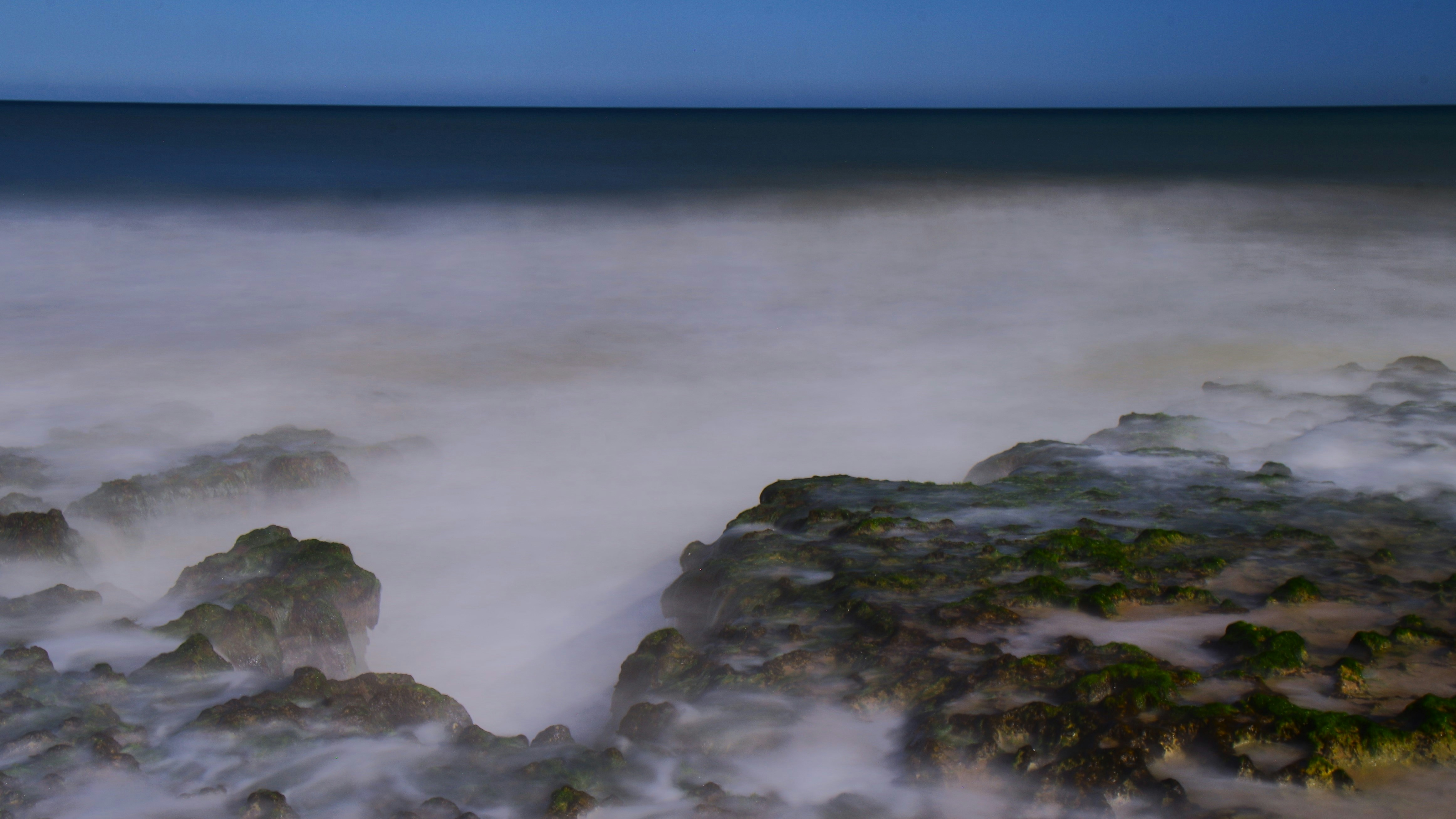 Long exposure of waves softly enveloping moss-covered rocks under a serene blue sky.