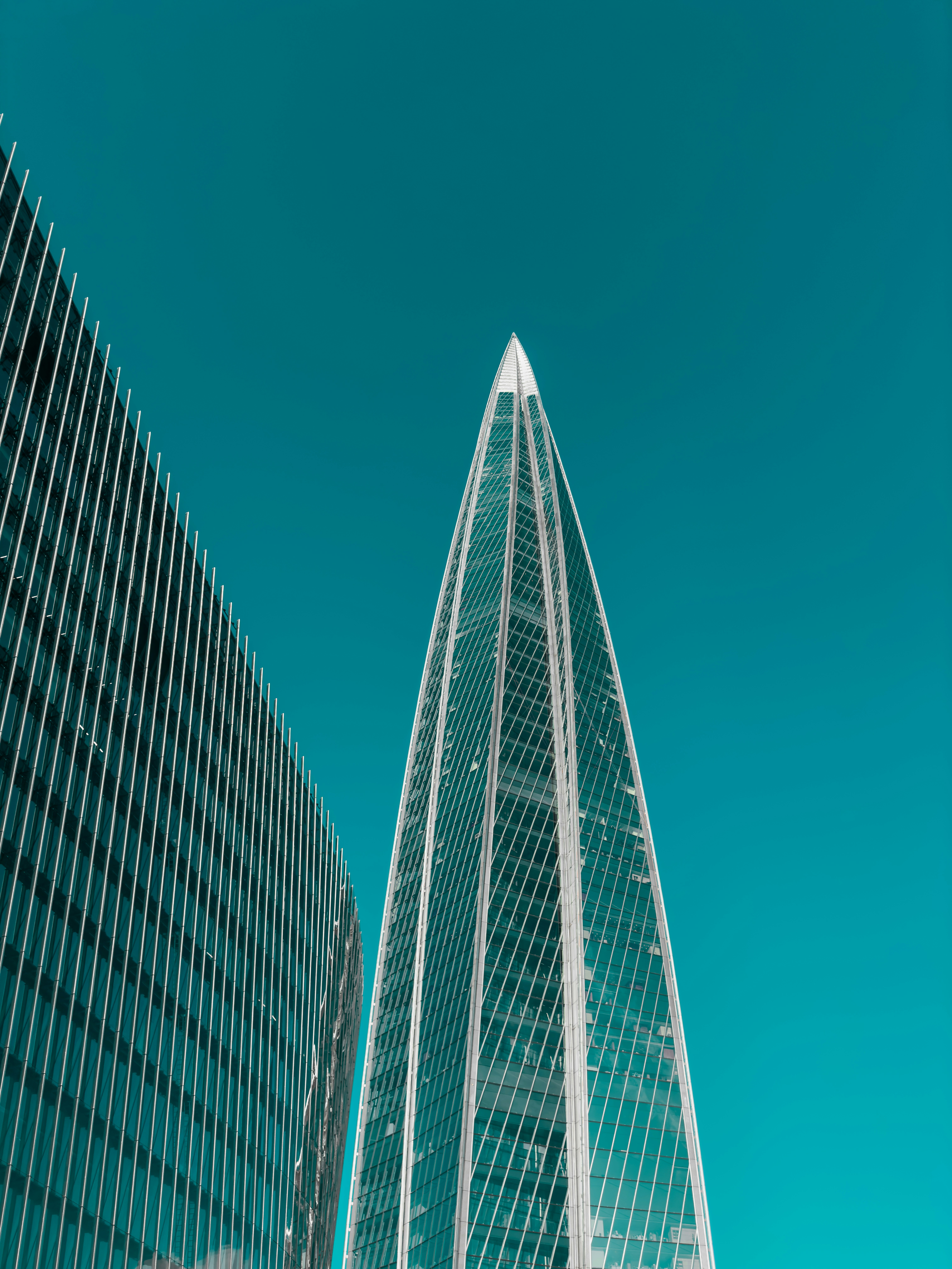 Photograph of a tall glass skyscraper with a pointed apex rising beside a curved, ribbed facade. The scene emphasizes architectural geometry against a teal sky.