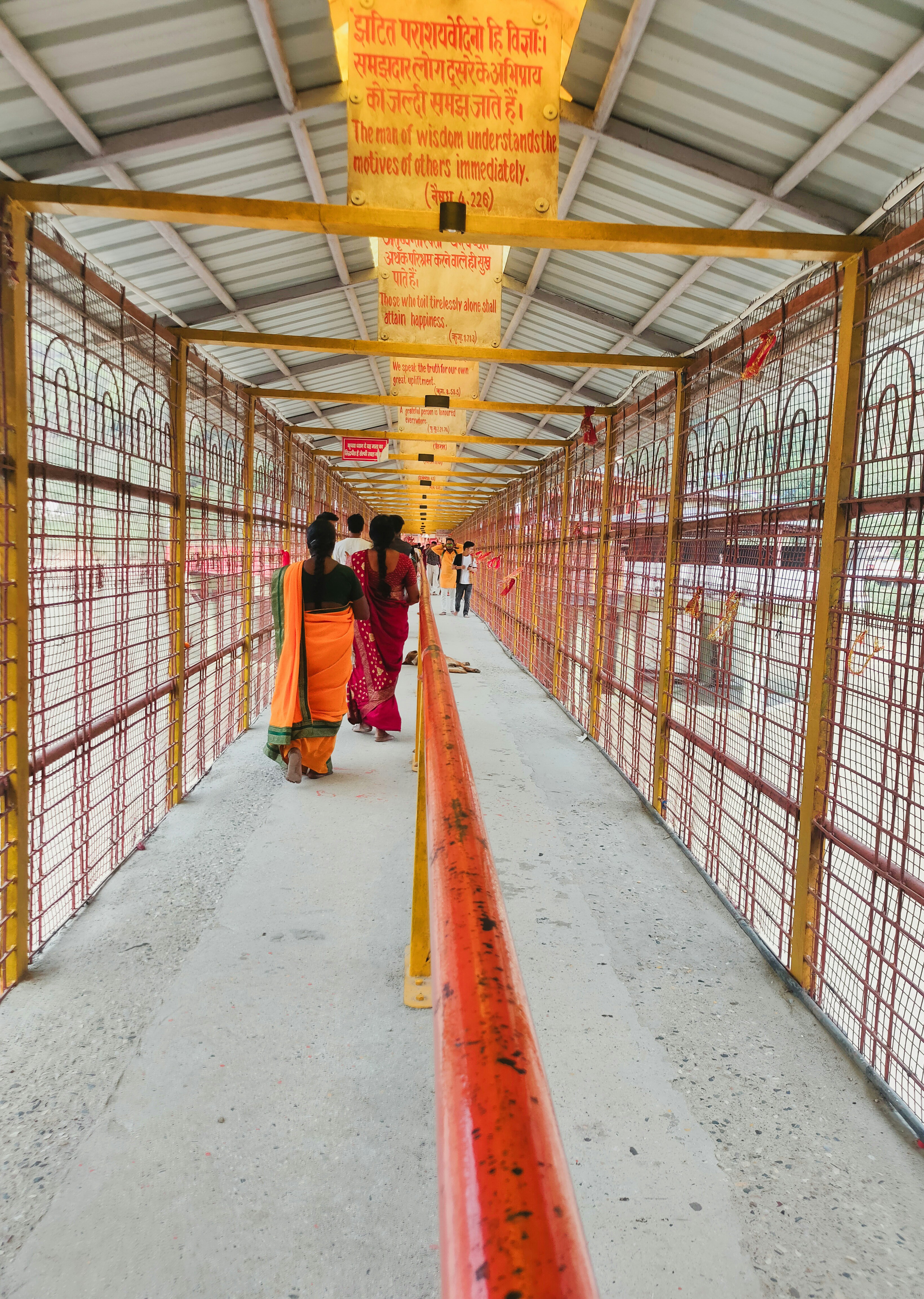 Colorful sari-clad pedestrians walk along a fenced, red-handled corridor toward a distant vanishing point beneath yellow banners.