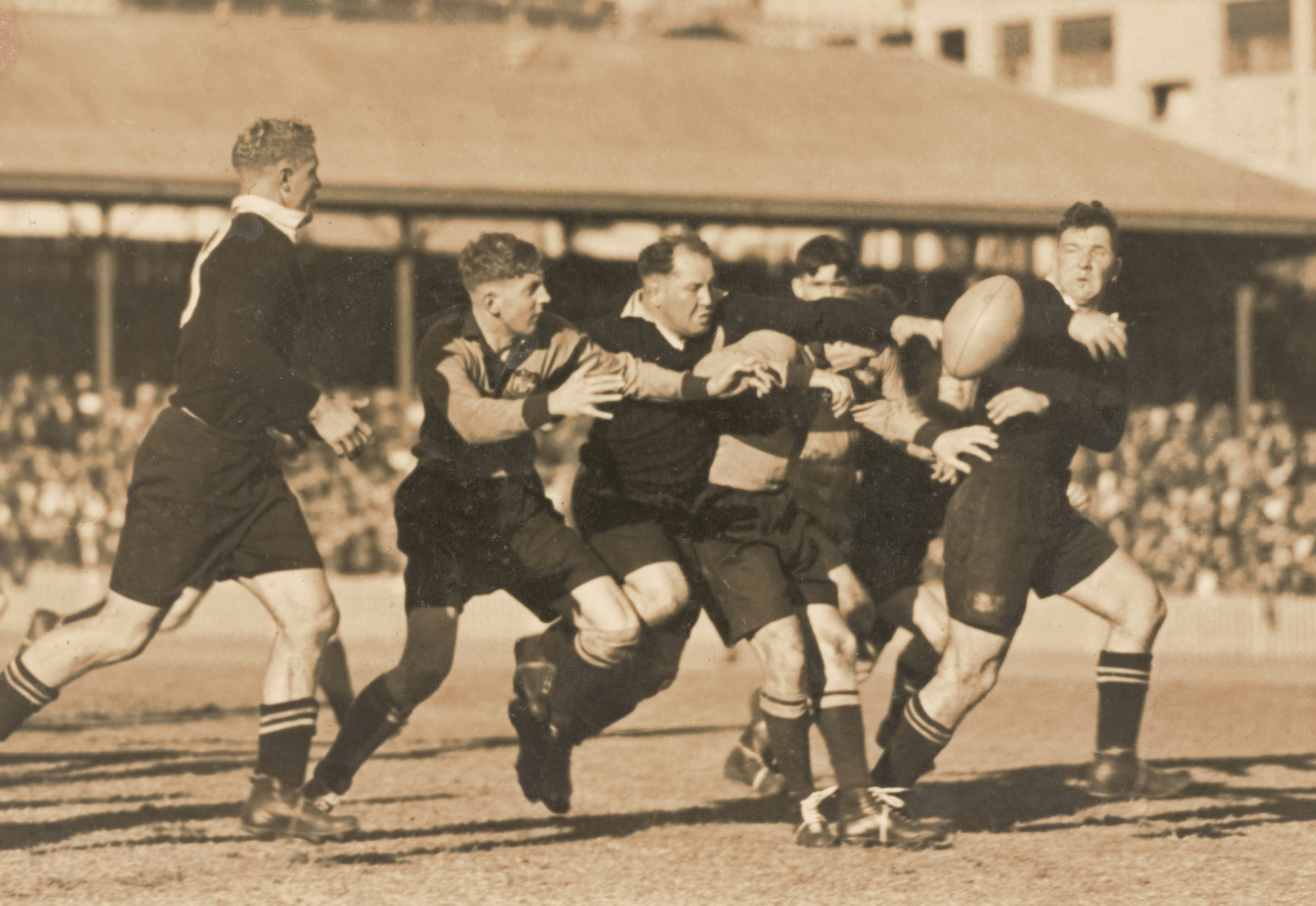 Rugby players in action during a match, captured in sepia tones, with a packed stadium in the background.