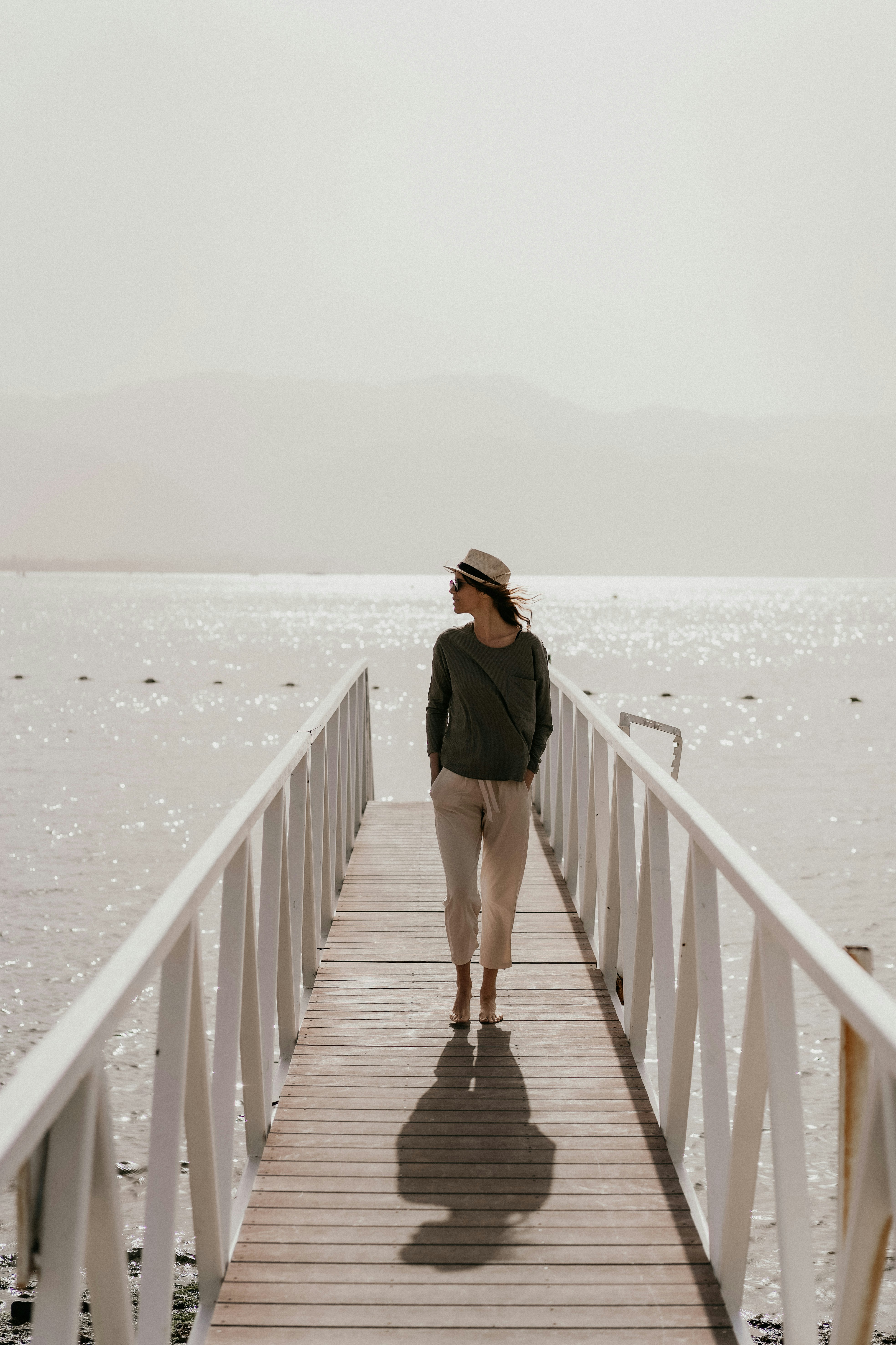 A woman strolls along a wooden pier, with shimmering water reflecting the soft light of a hazy day.