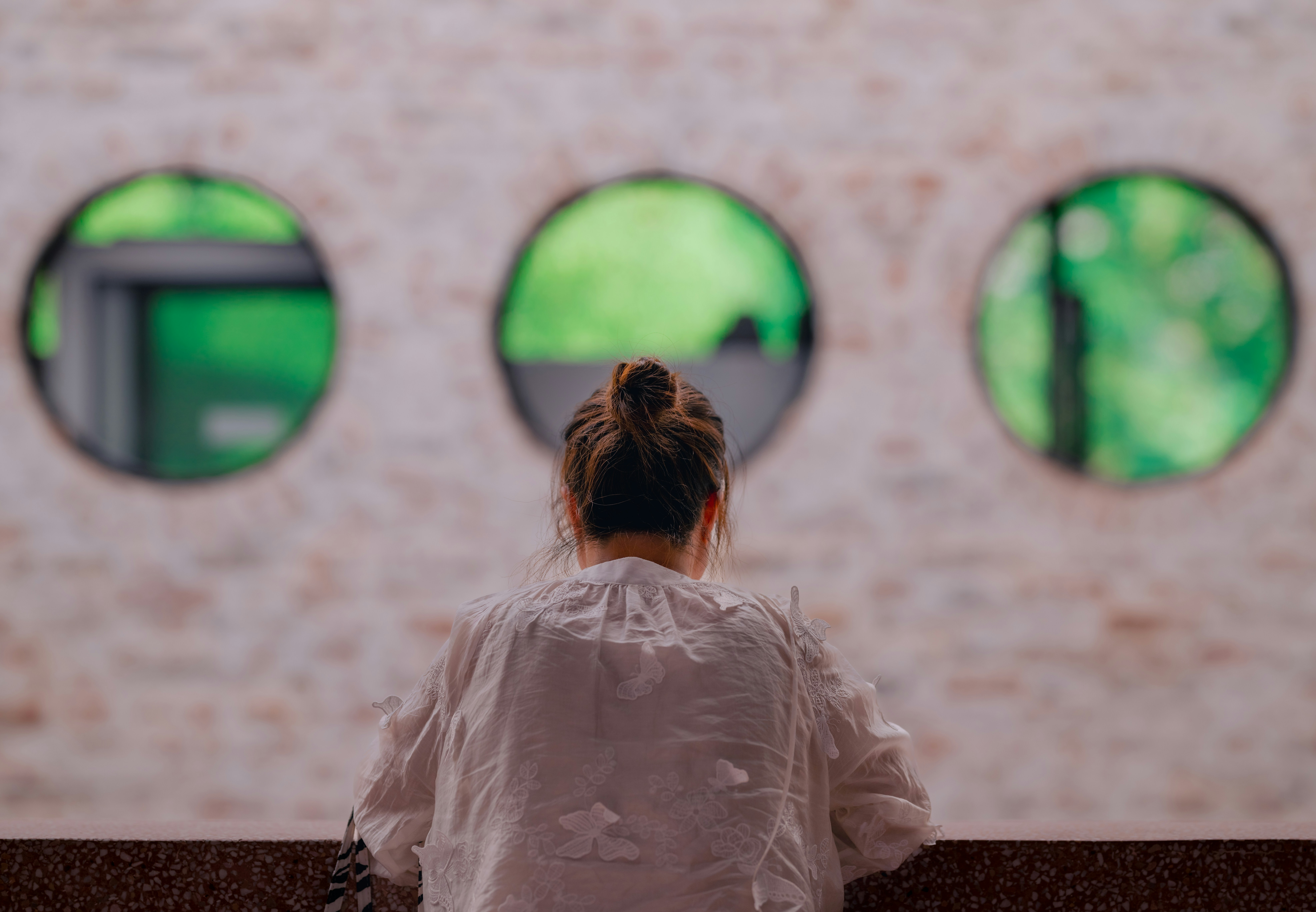 Portrait of a person seen from behind, with a light blouse and hair in a bun, standing before a brick wall. Three circular windows behind capture green reflections, adding a geometric counterpoint to the scene.