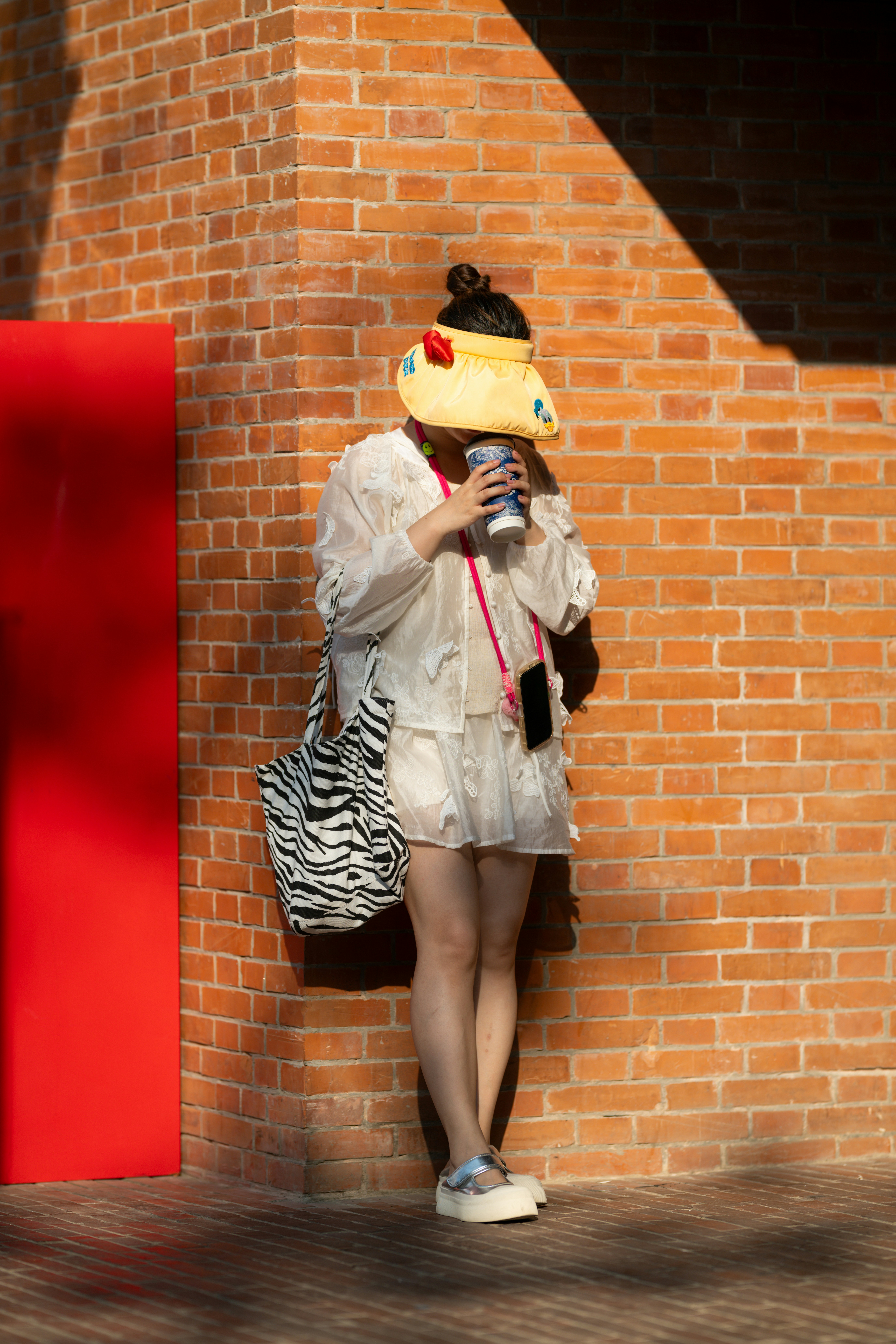 A person stands against a sunlit brick wall, sipping from a cup while wearing a yellow cushion hat and a sheer white outfit. A zebra-striped tote and pink crossbody strap complete the urban look.