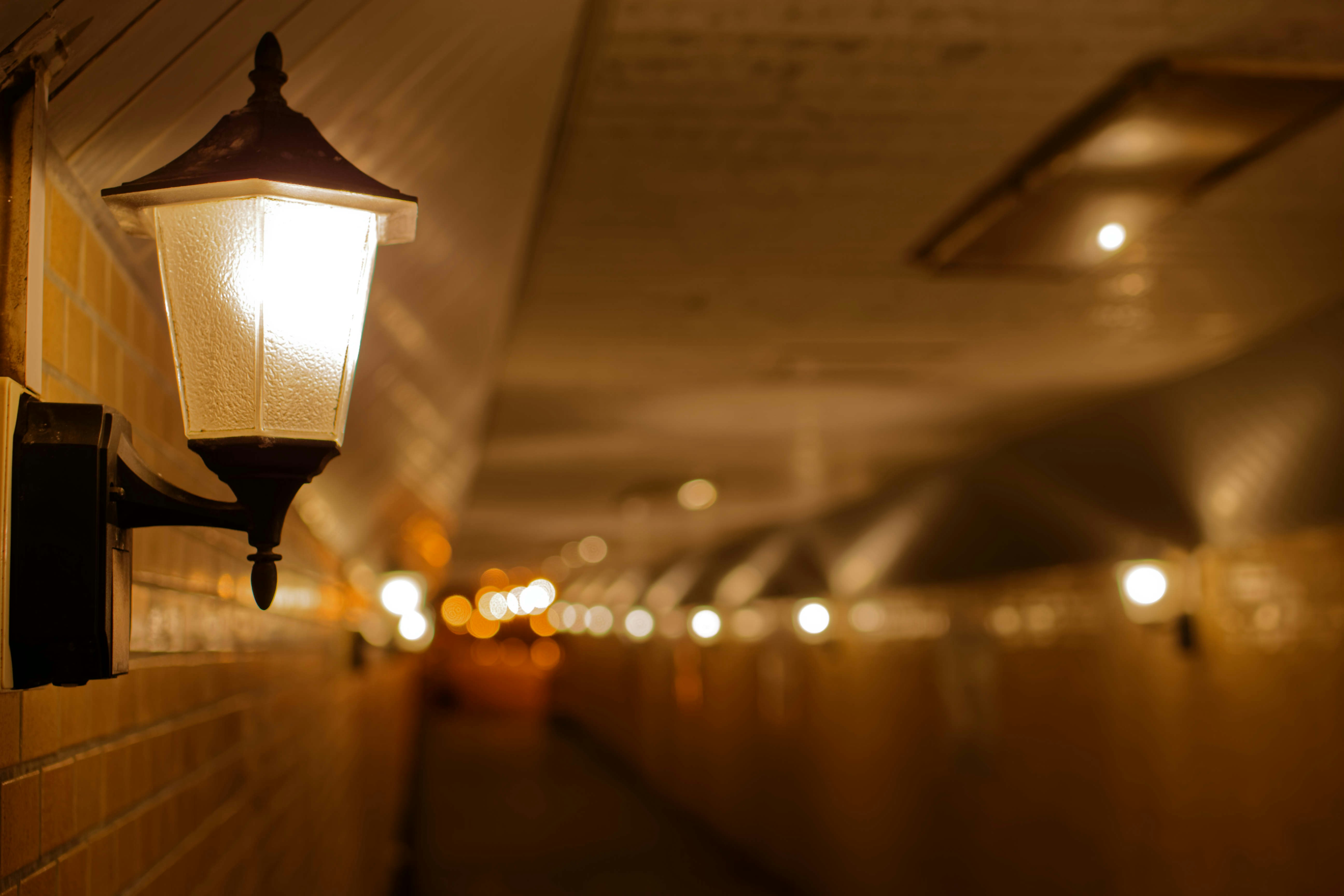 Warm wall lantern anchors the foreground against a brick tunnel. The corridor recedes into soft, glowing bokeh.