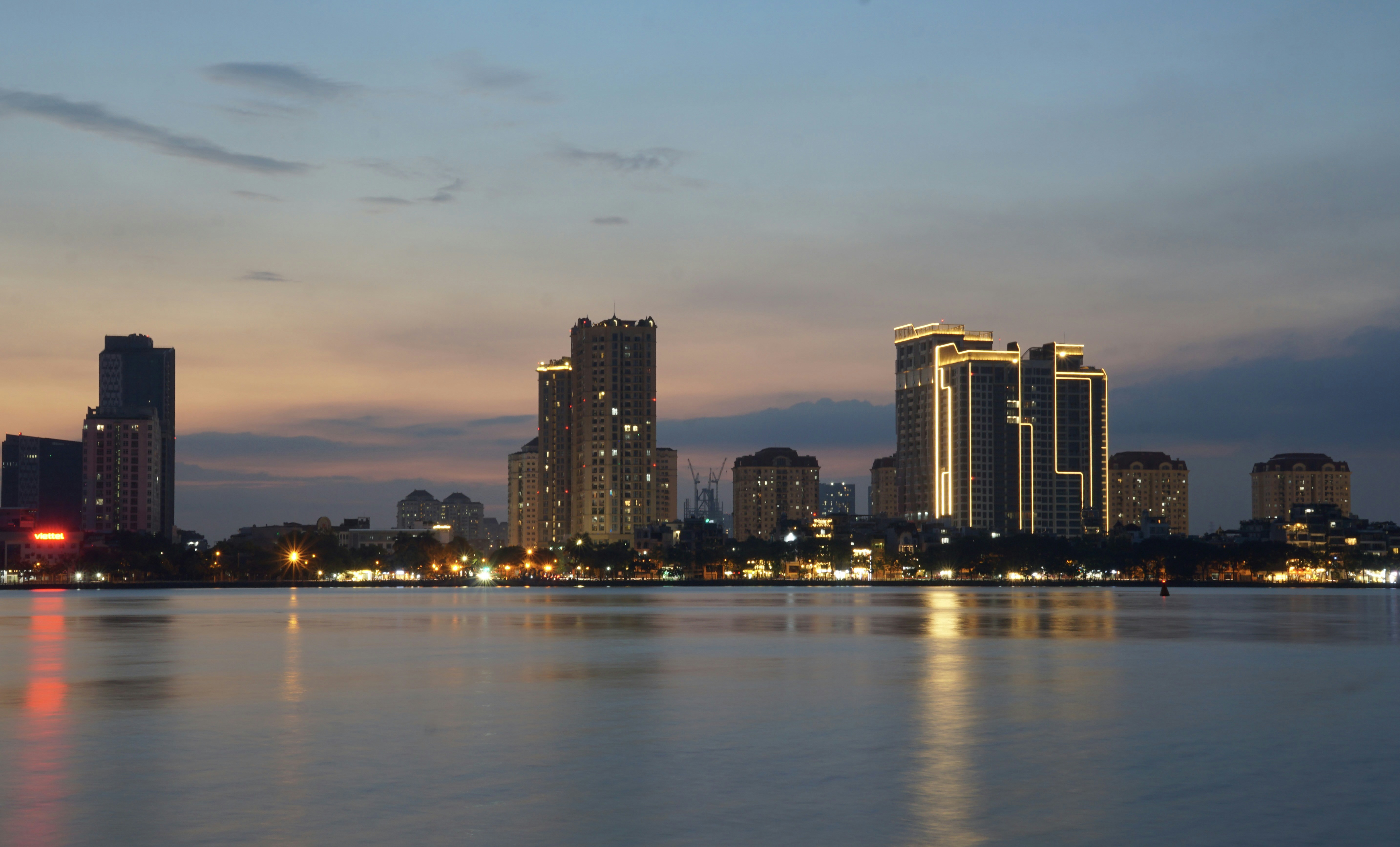 Calm waters reflecting a city skyline at dusk with buildings softly illuminated against a darkening sky.