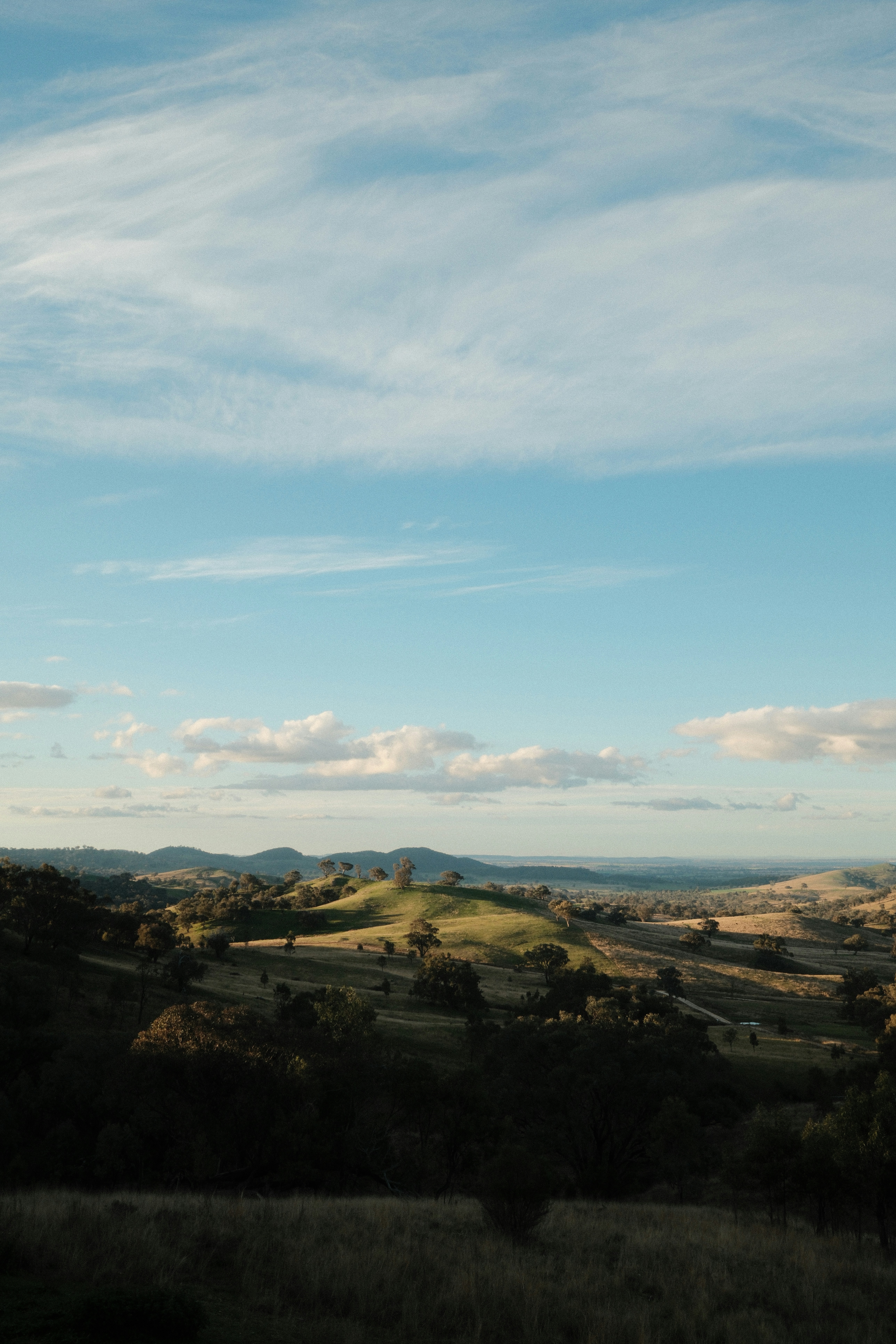 Sunlit rolling hills spread across a quiet valley beneath a pale-blue sky with wispy clouds.