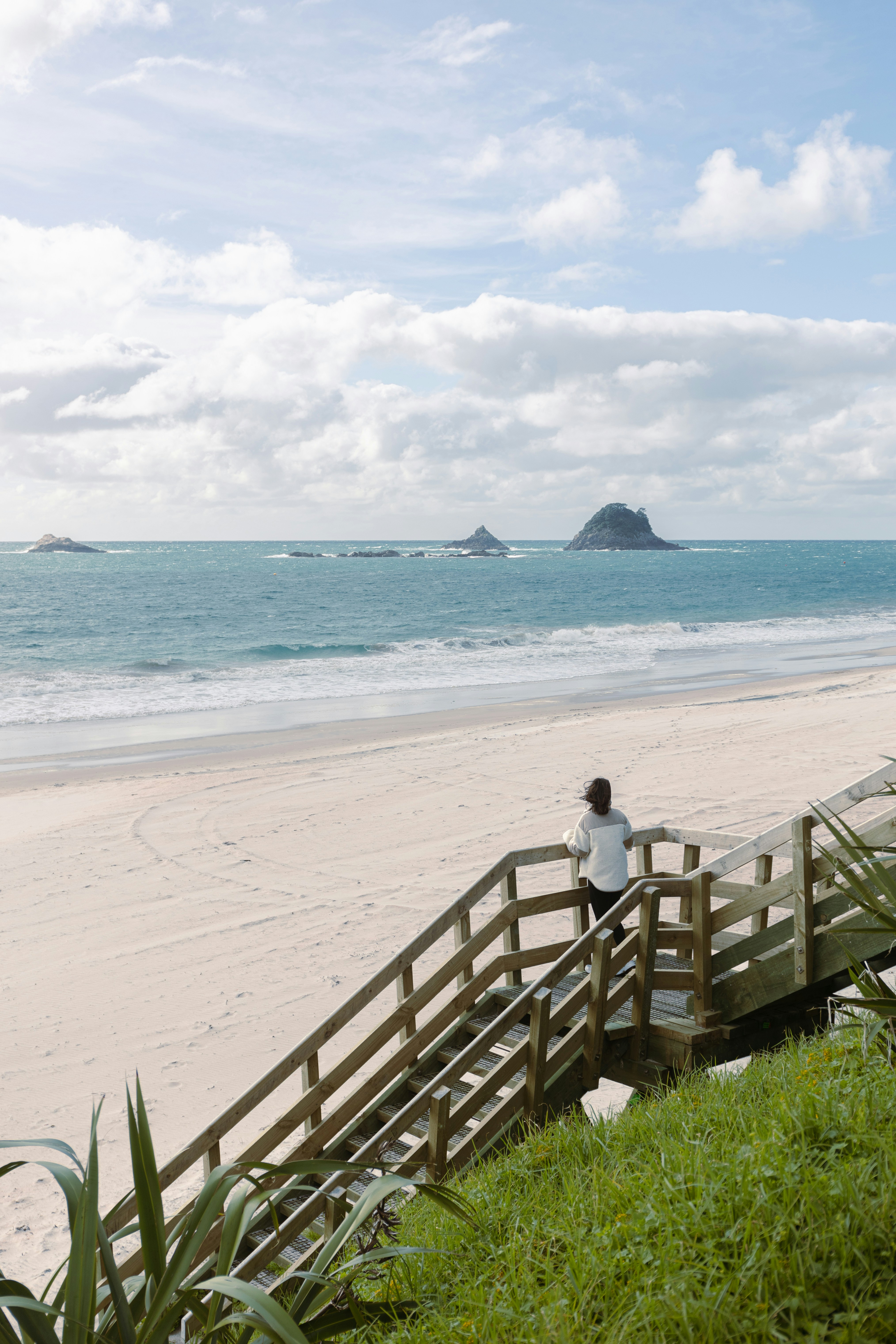 Person gazing at ocean waves from a wooden stairway on a sandy beach under a partly cloudy sky.