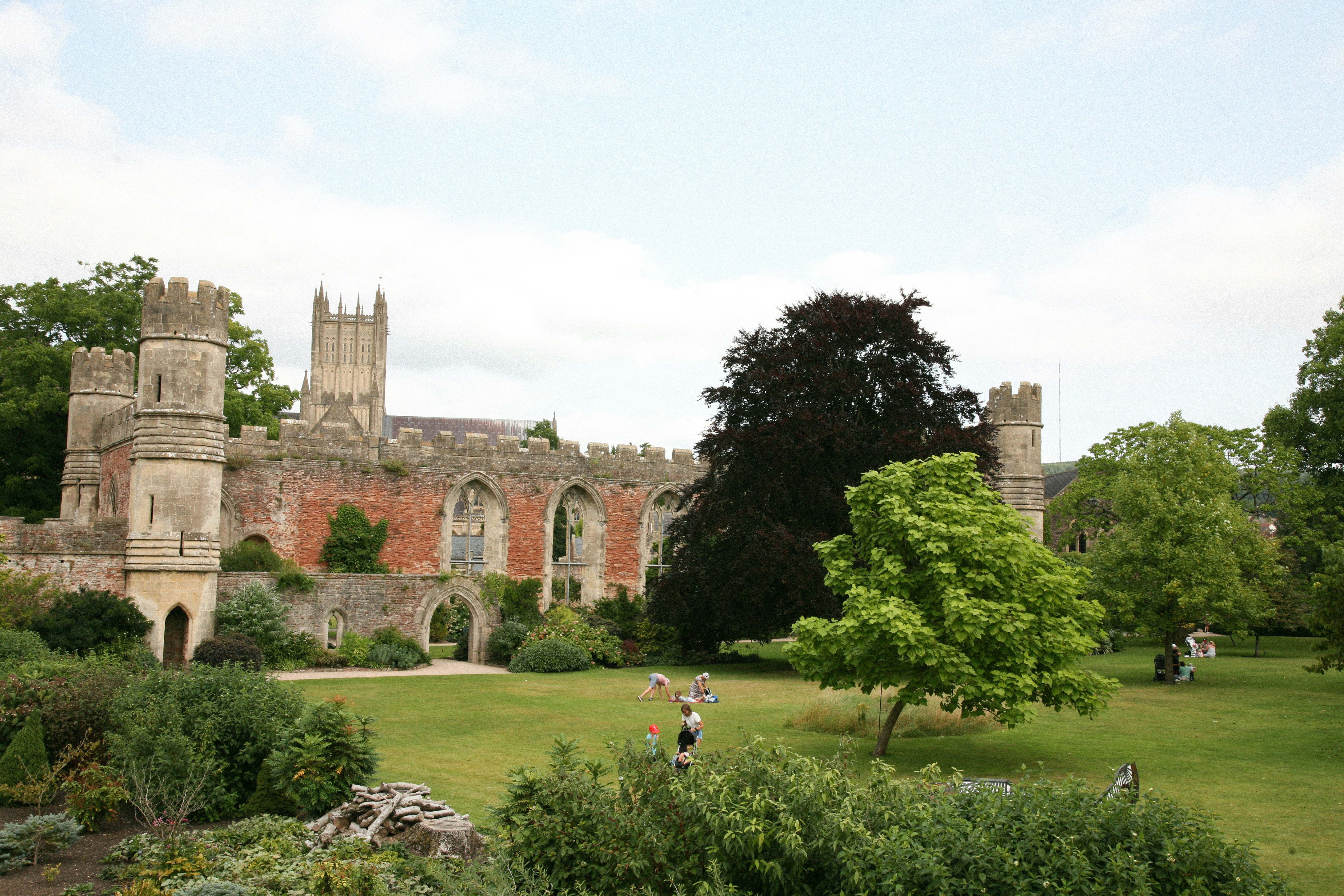 This photo depicts a historic stone building surrounded by lush green gardens. A large, imposing structure with towers and arches dominates the center of the image. In the foreground, there is a manicured lawn with people enjoying the outdoors. The sky is partly cloudy.