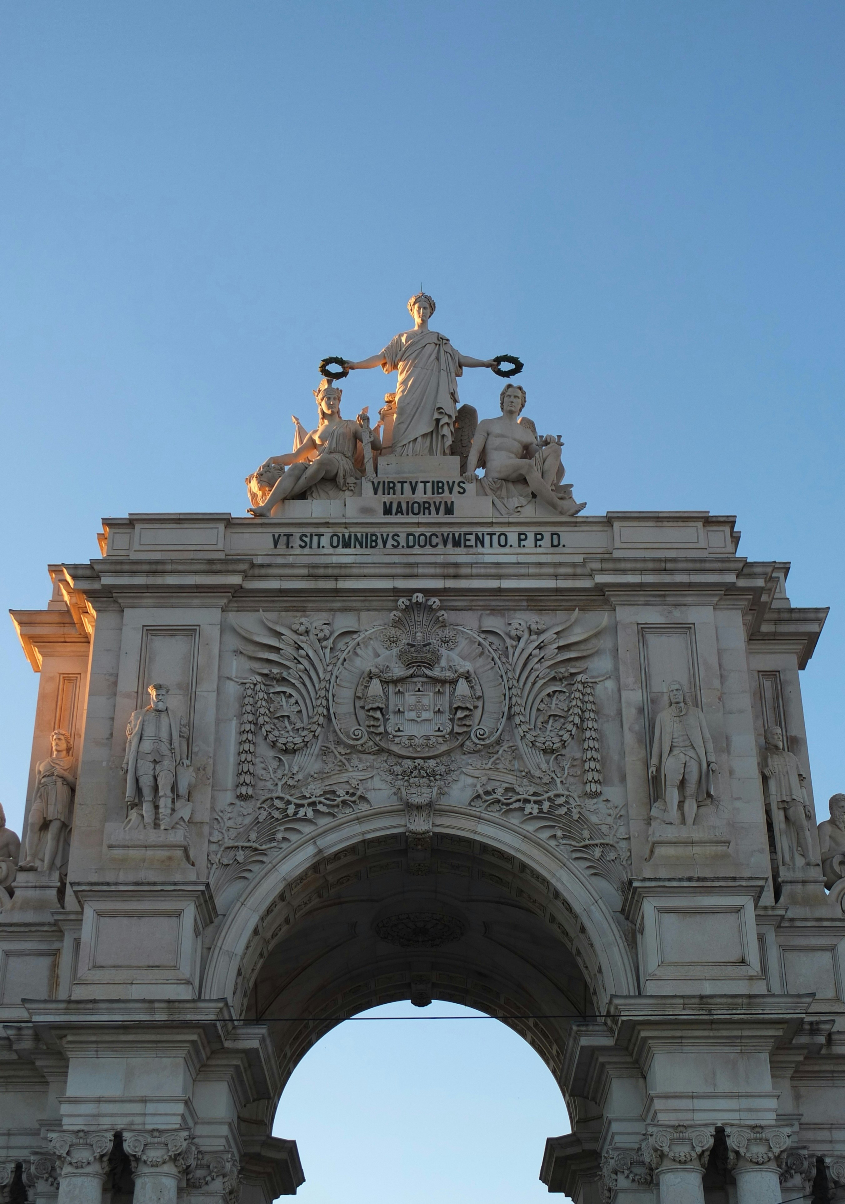 A grand stone arch adorned with sculpted figures and intricate bas-relief, topped by a classical sculpture group against a clear blue sky.