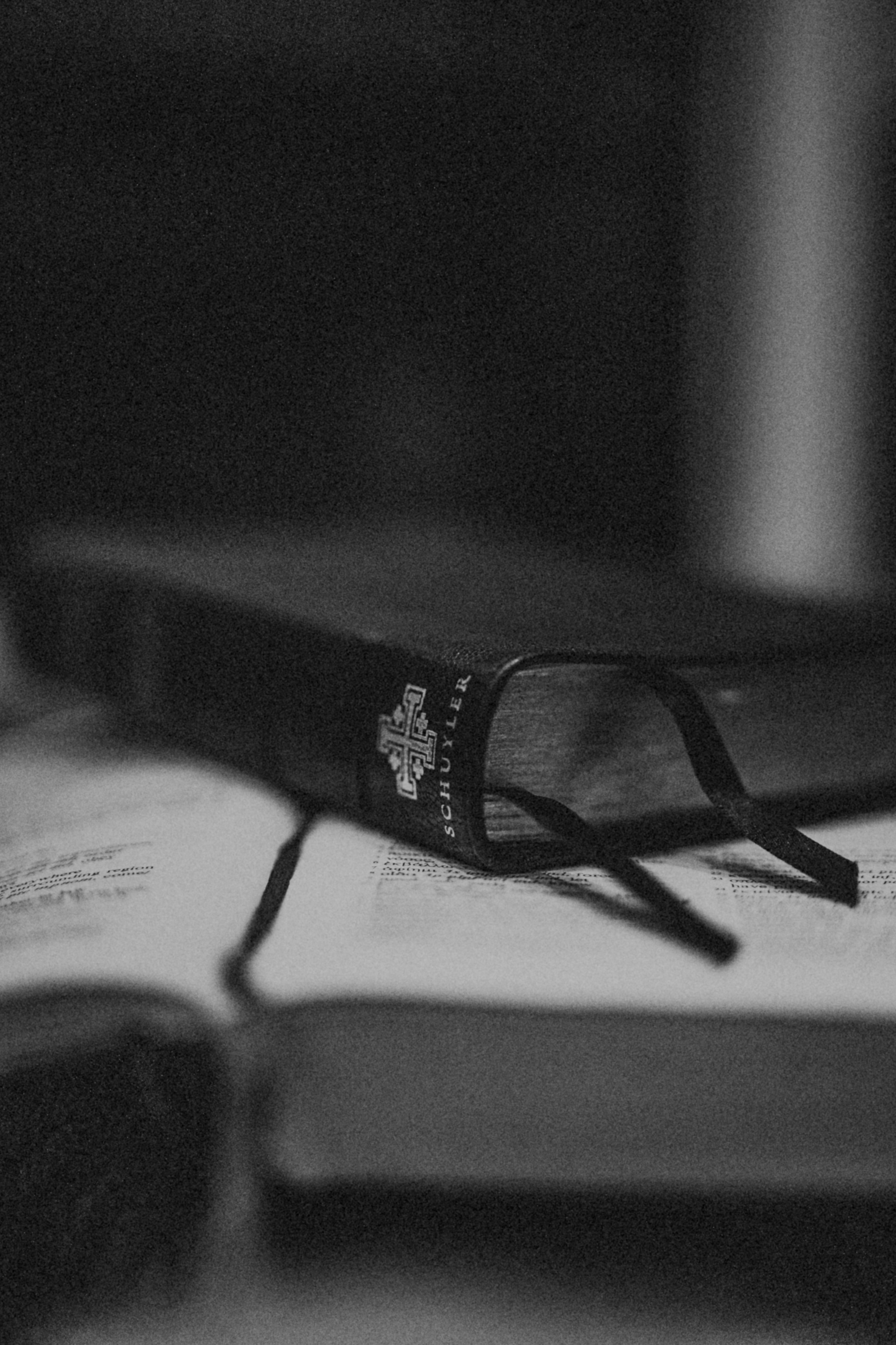 A black-and-white close-up of a Bible with a ribbon bookmark resting on open pages; a subtle glow highlights the spine where a cross emblem appears, suggesting a quiet, contemplative moment.