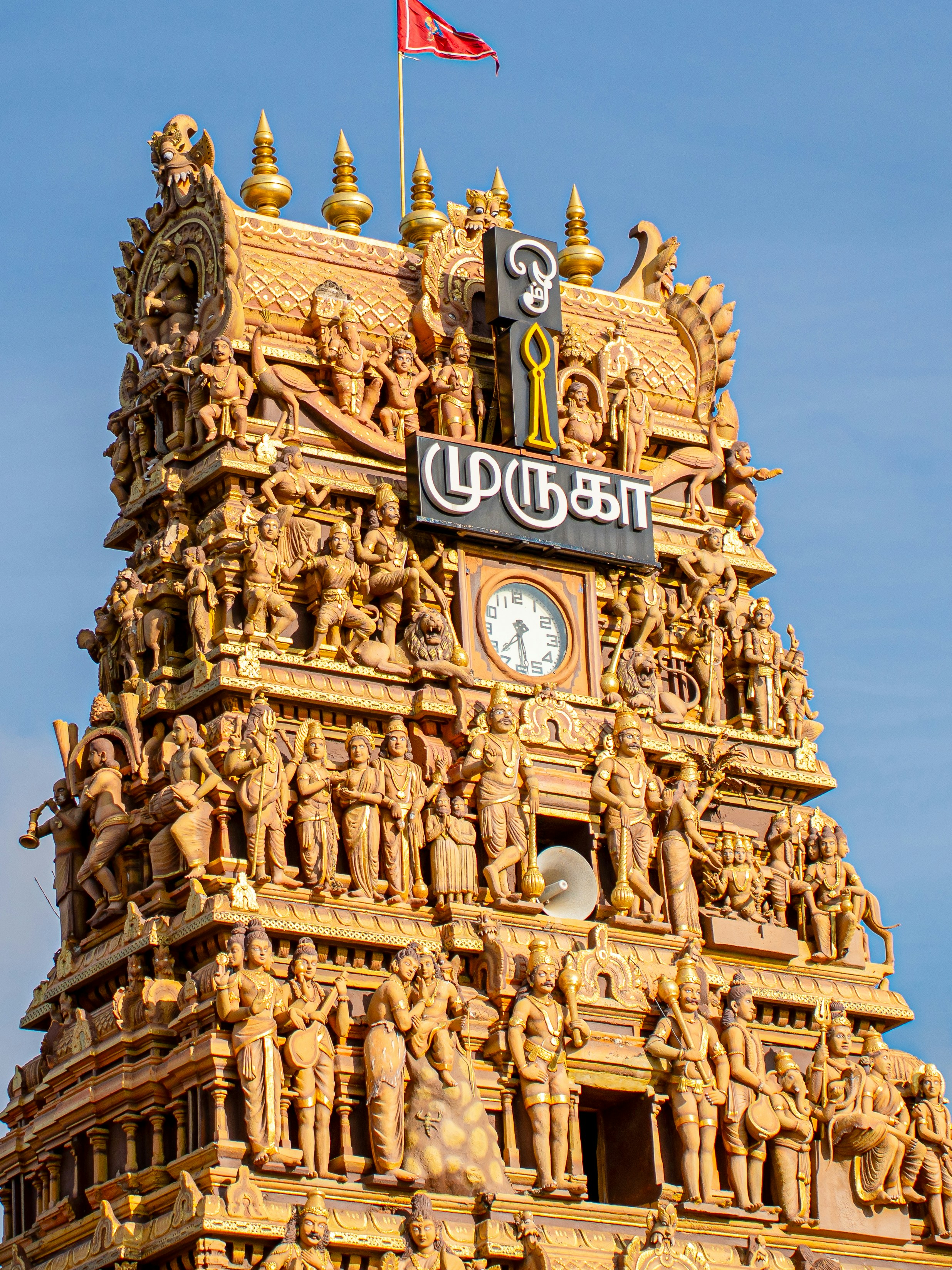 A photograph of a gilded temple tower adorned with intricate statues and a central clock, set against a clear blue sky.