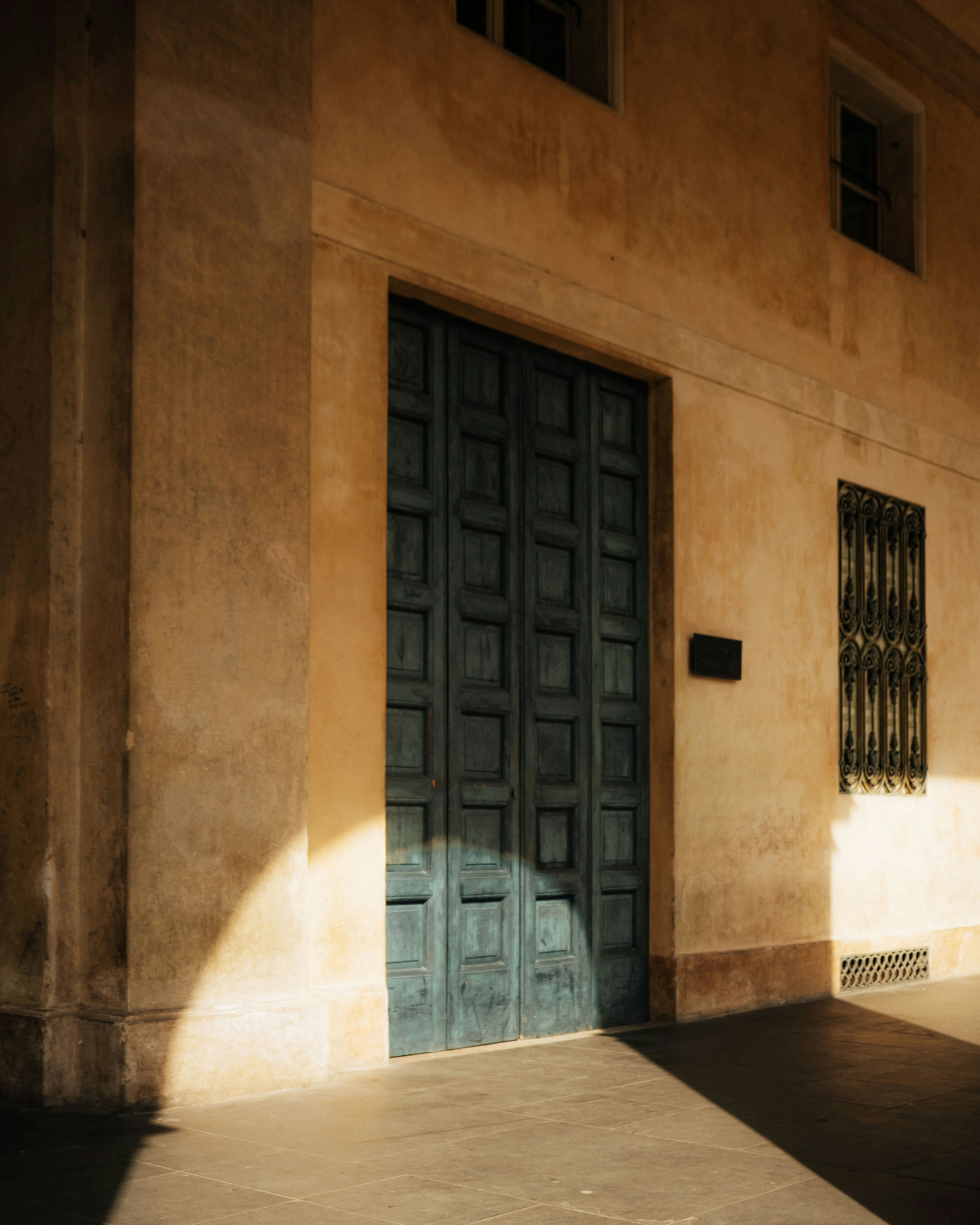 A large green door partially illuminated by sunlight on a textured wall with a small window and a shadowed plaque.