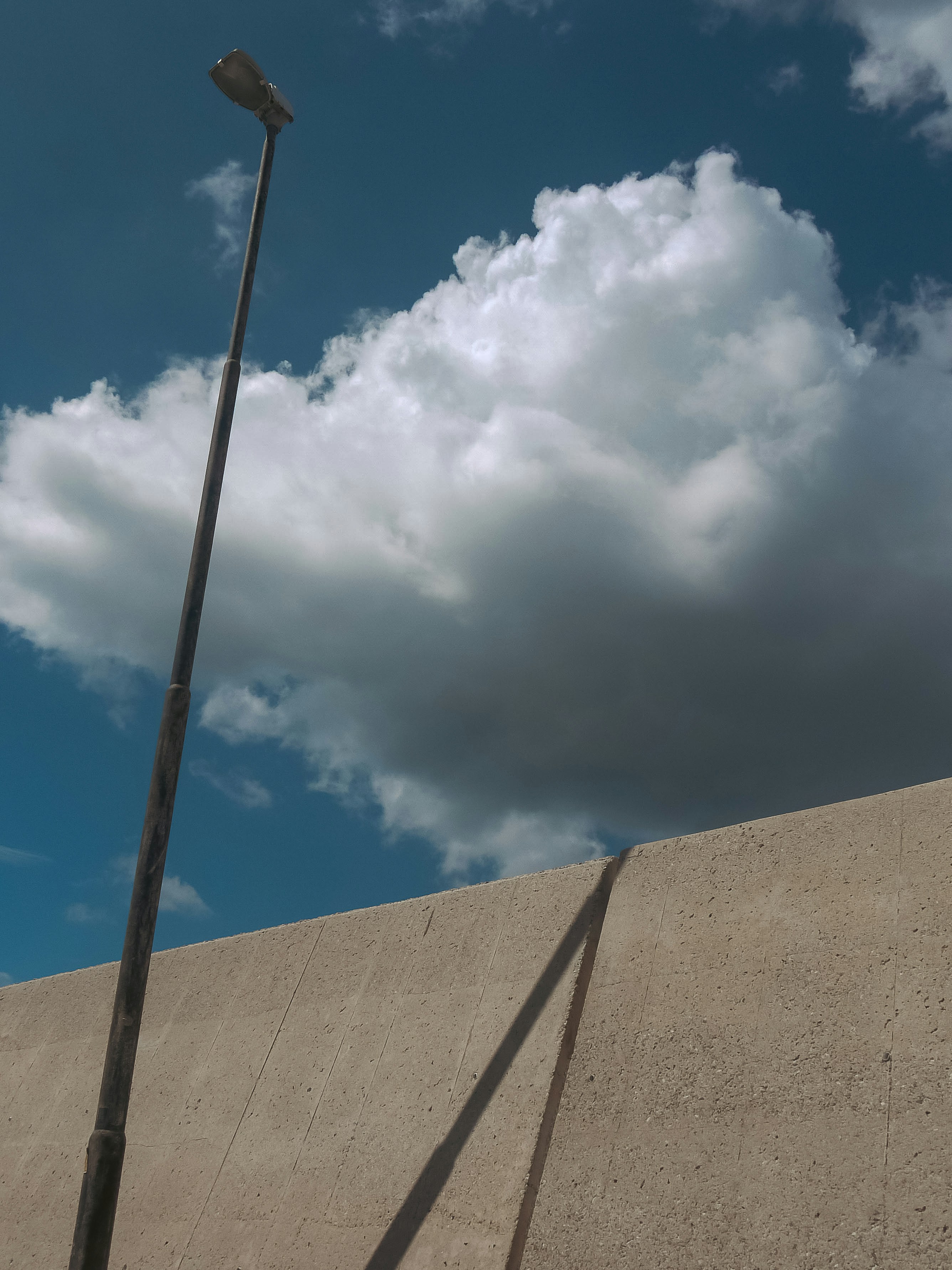 A photograph capturing a tall streetlamp against a blue sky with large clouds, casting a long shadow on a sloped concrete wall.
