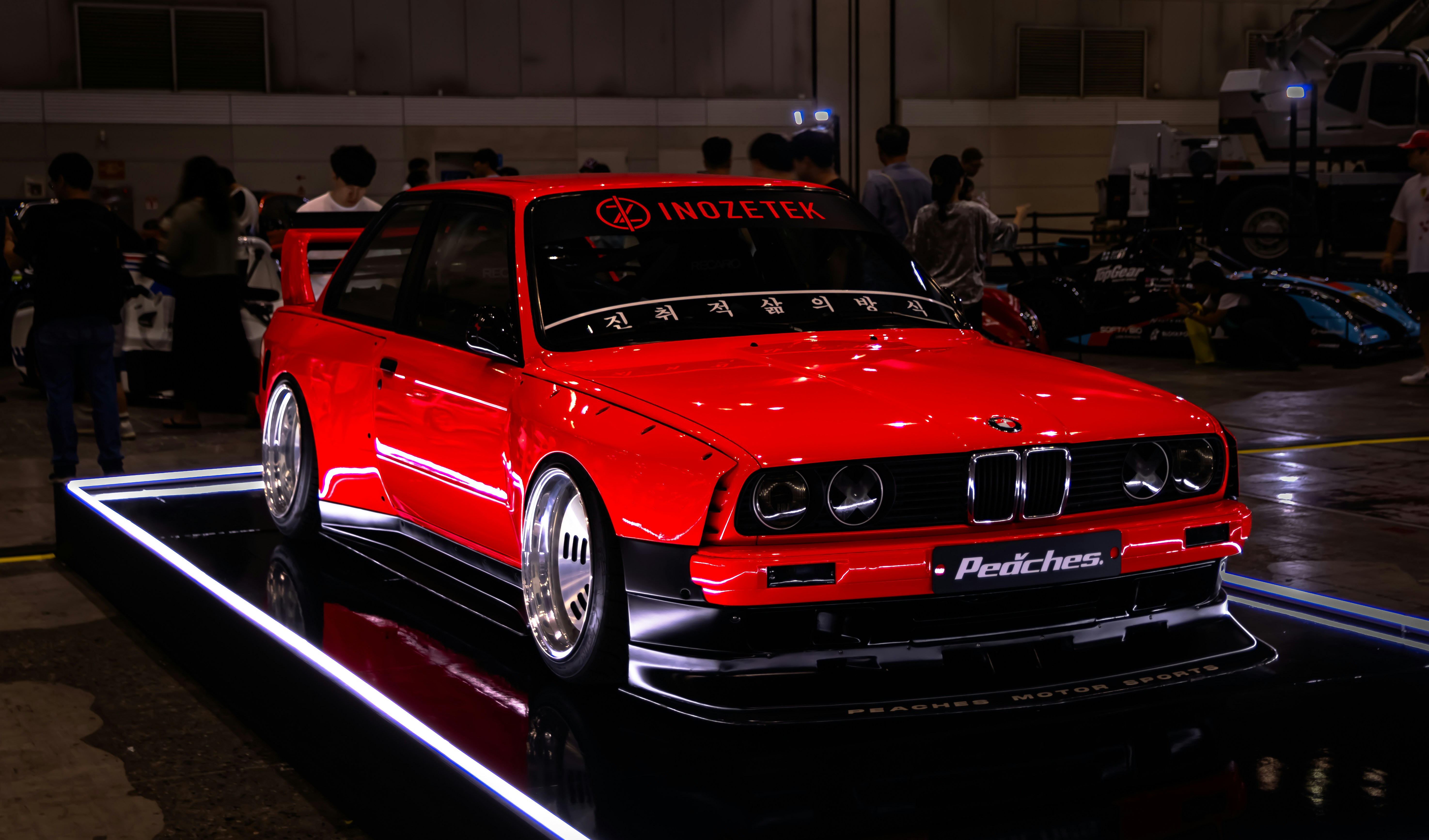 A modified red BMW E30 showcased on a sleek platform, illuminated by vibrant lighting at an automotive exhibition.