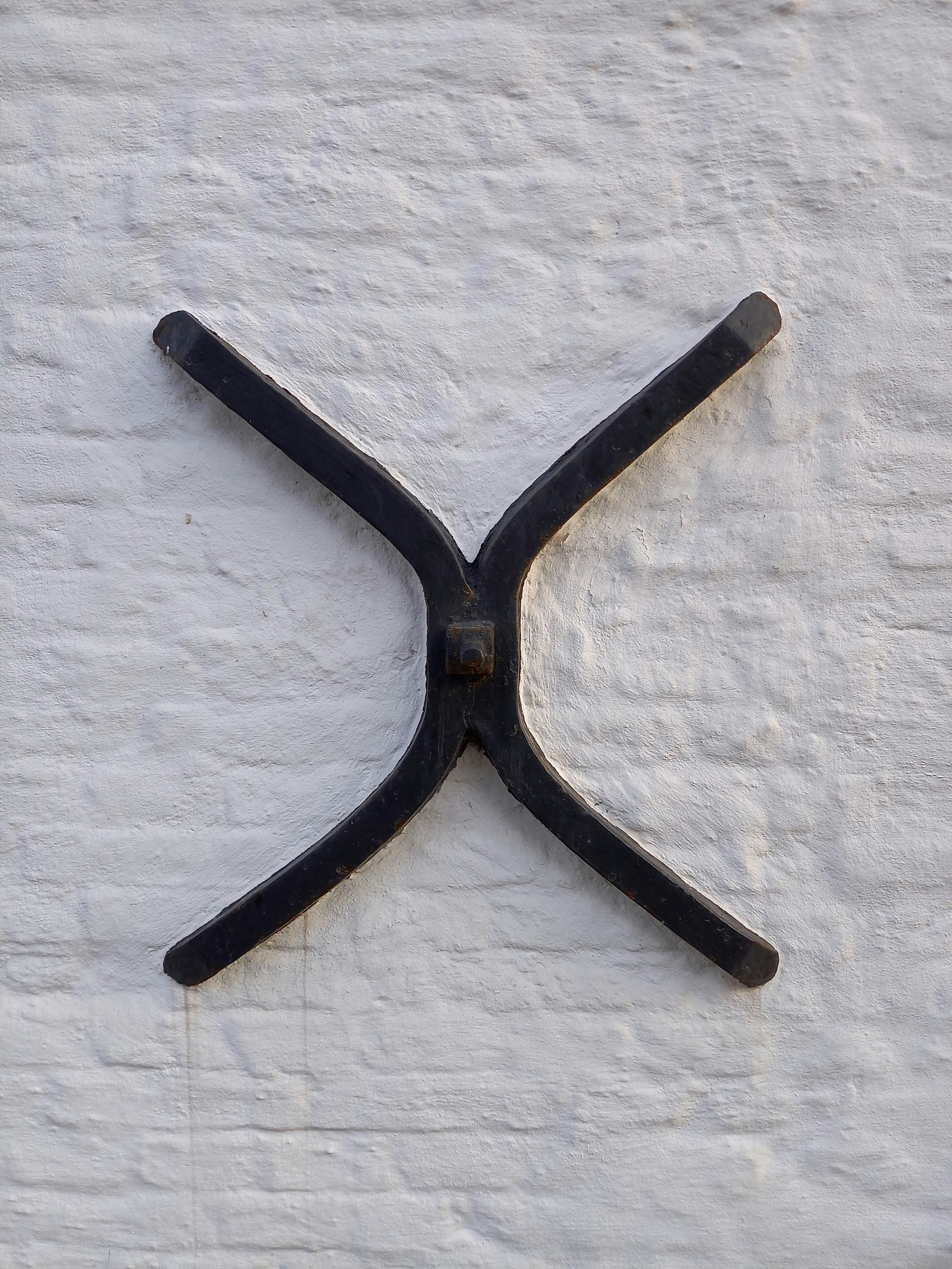 X-shaped metal bracket mounted on a white textured plaster wall, its four curved arms converging at a central joint.