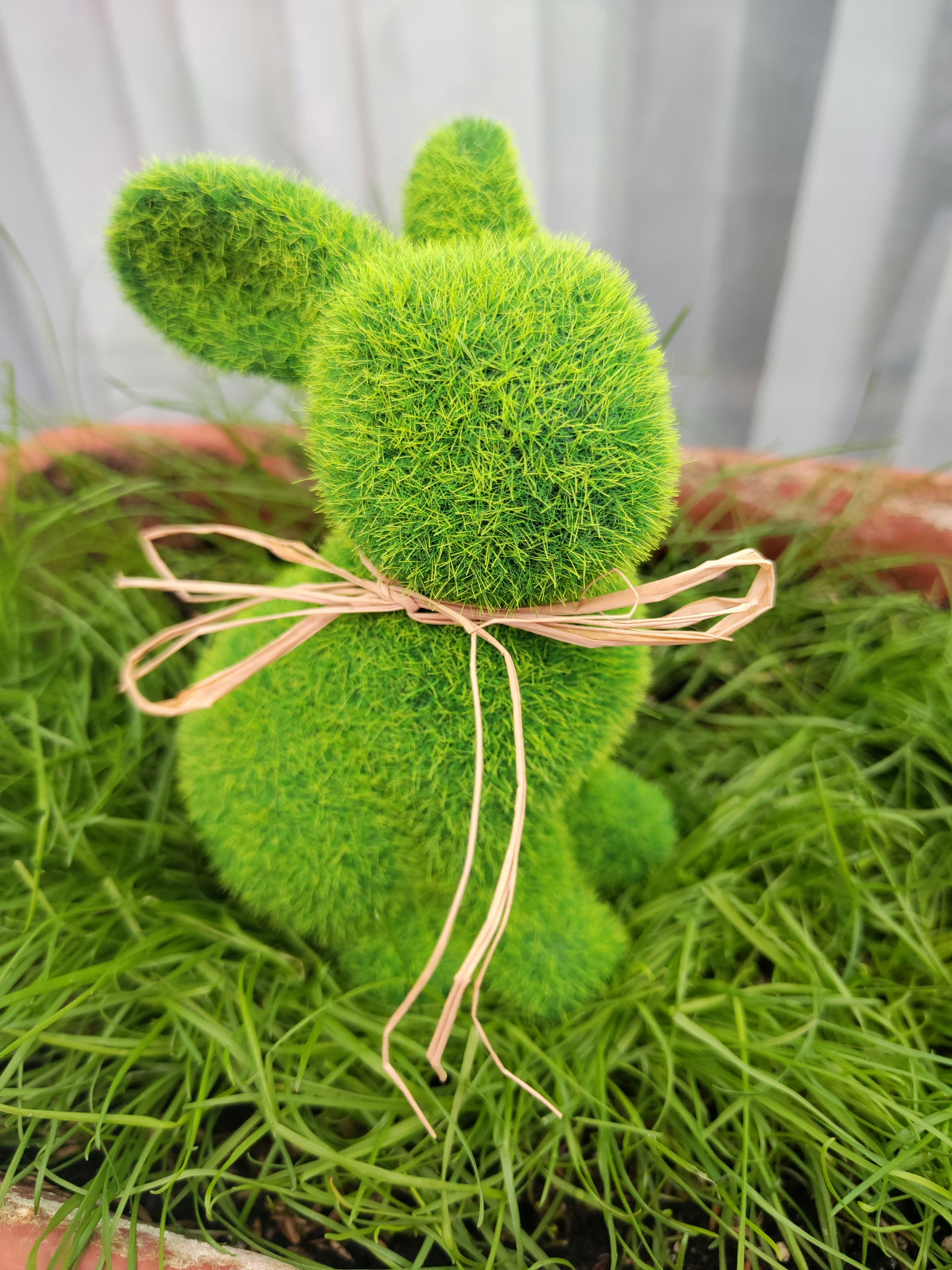 Verdant moss bunny sculpture in a pot, tied with a raffia bow and nestled among green grass.