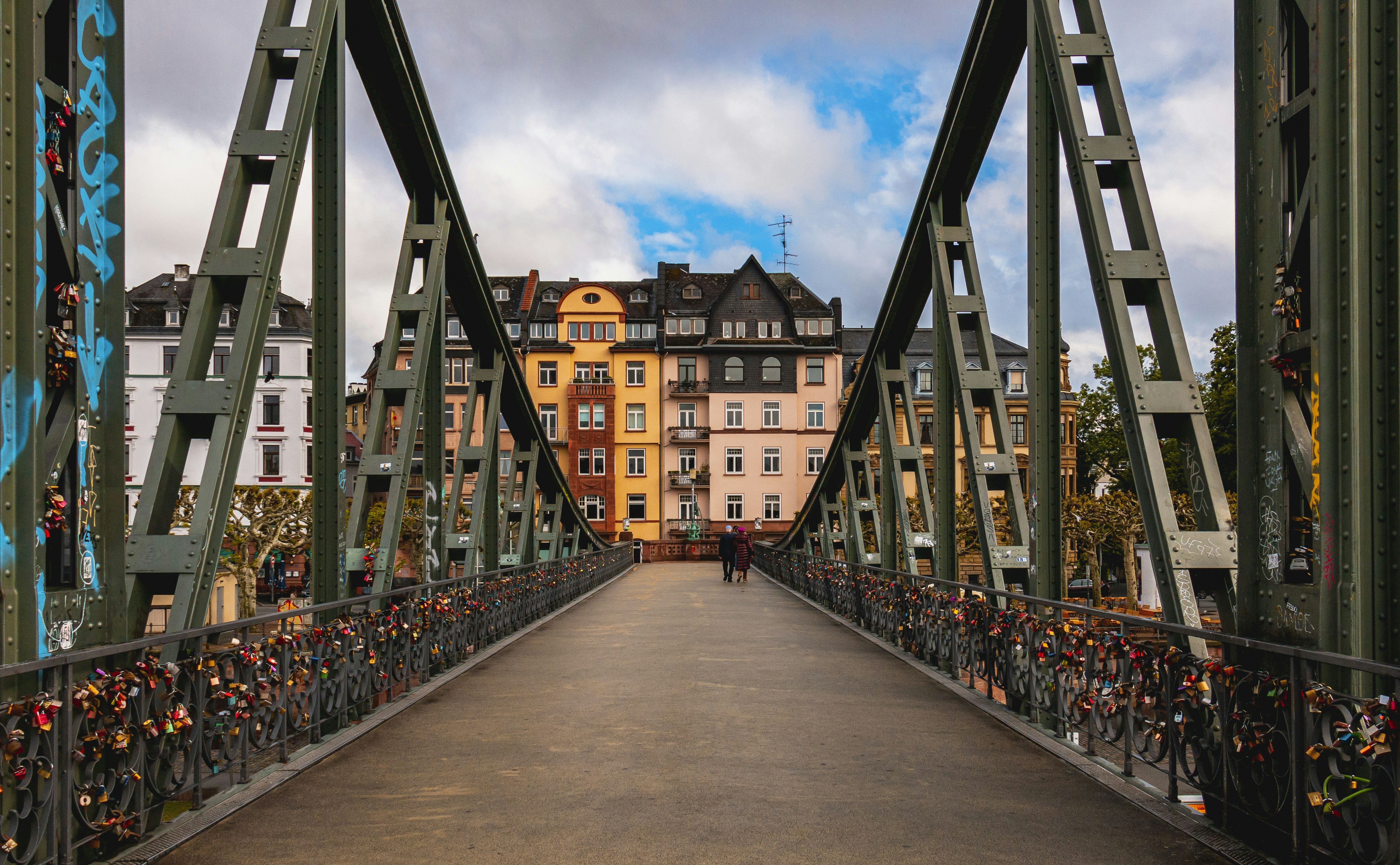 A vibrant urban bridge adorned with love locks, leading to colorful buildings under a cloudy sky. The scene captures a blend of romance and architectural beauty.