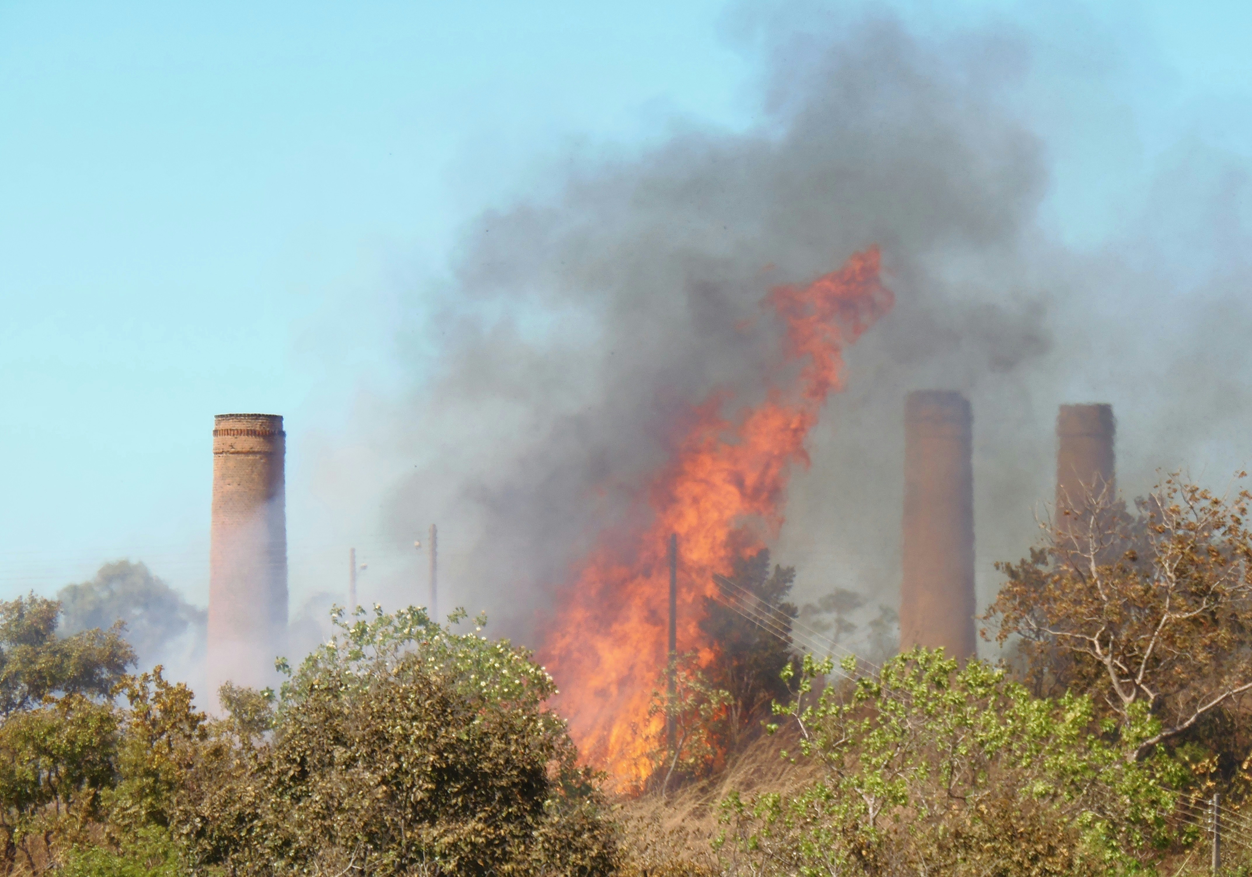 Active factory fire with tall flames and smoke behind vegetation; industrial smokestacks visible in the background, conveying an urgent scene.