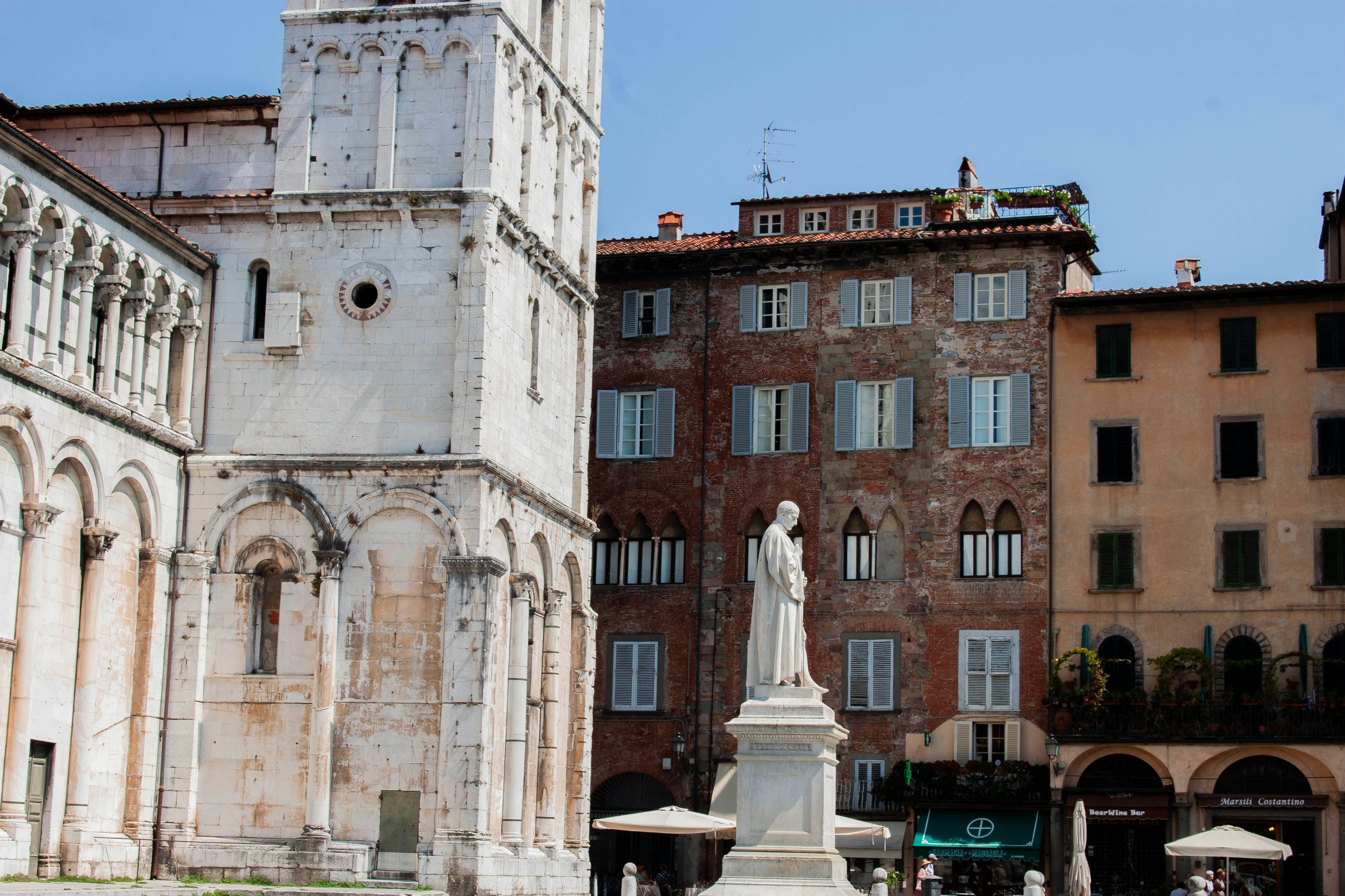 White clock tower and statue beside historic buildings under a clear blue sky.